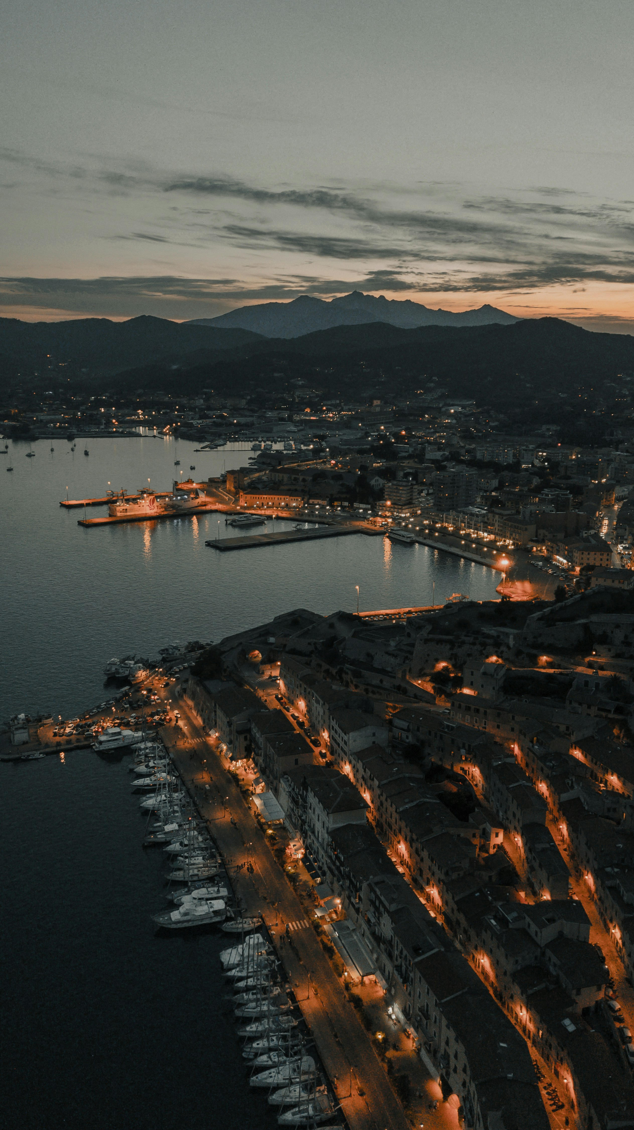 Aerial view of a coastal town at twilight, showcasing illuminated buildings and boats along the harbor. The backdrop features distant mountains under a soft dusk sky.