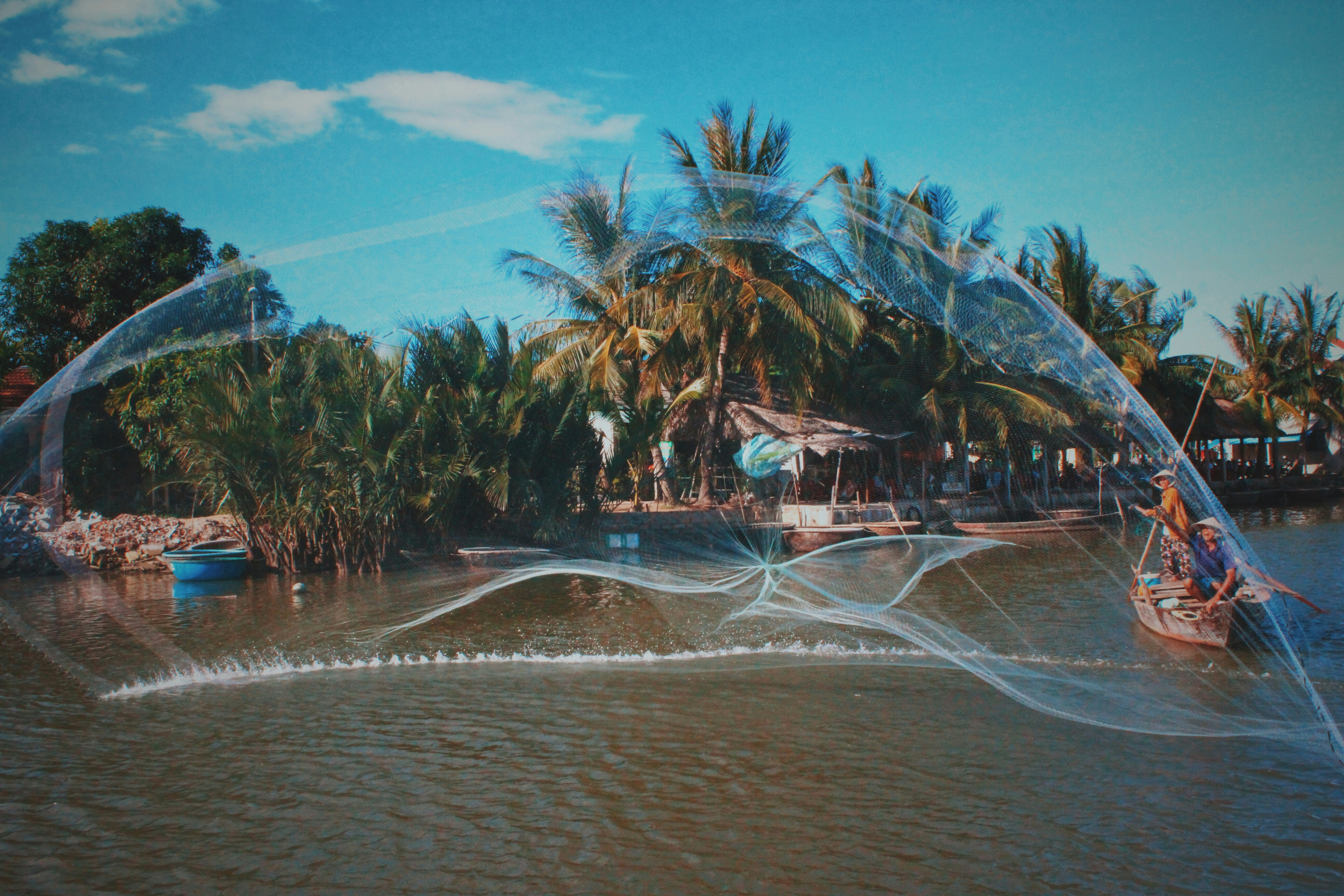 Fisherman casting a net over a tranquil river, surrounded by lush palm trees and a rustic fishing village in the background.