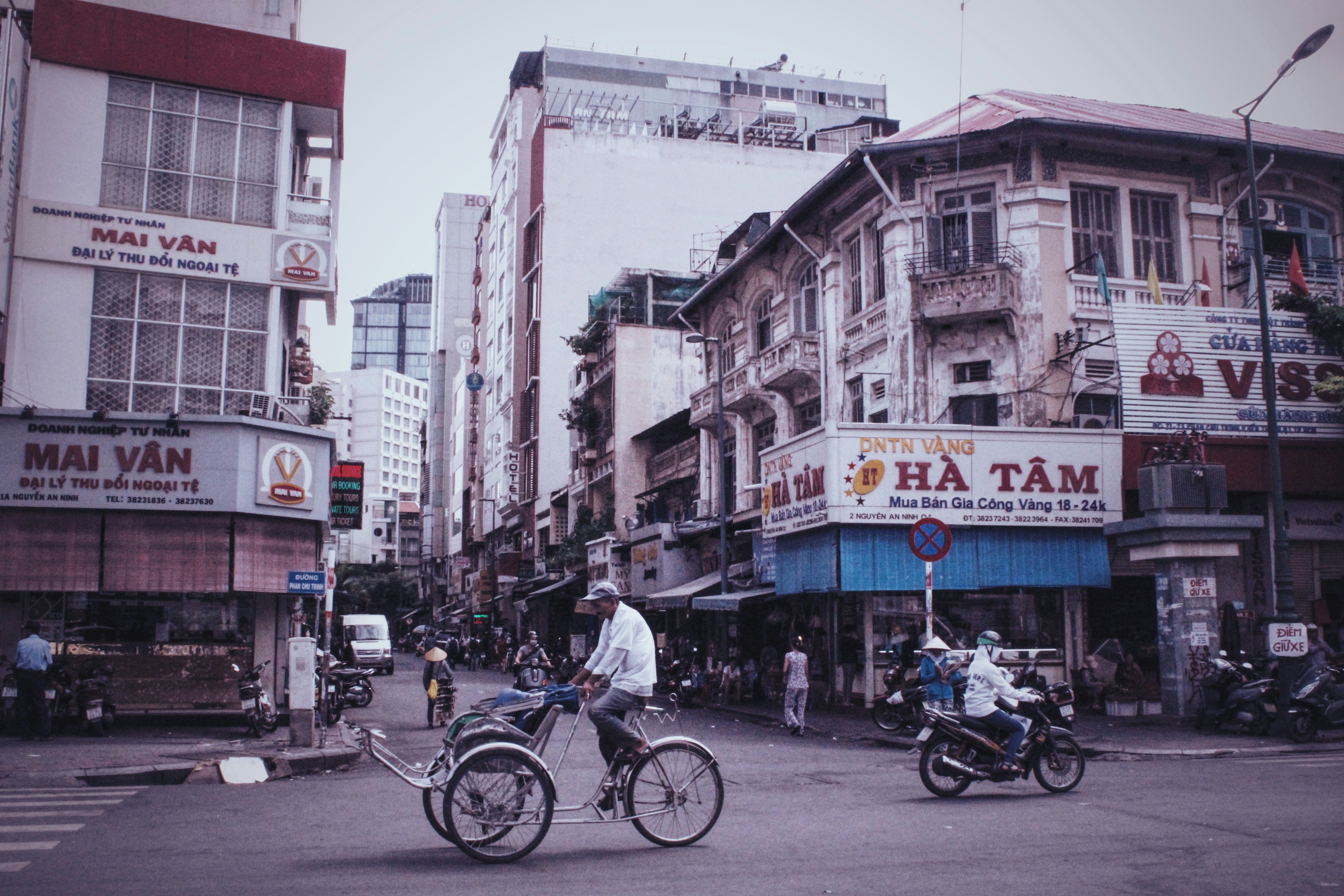 A bustling street scene in a vibrant city, featuring a cyclo rider navigating through traffic amidst colorful storefronts and architecture.