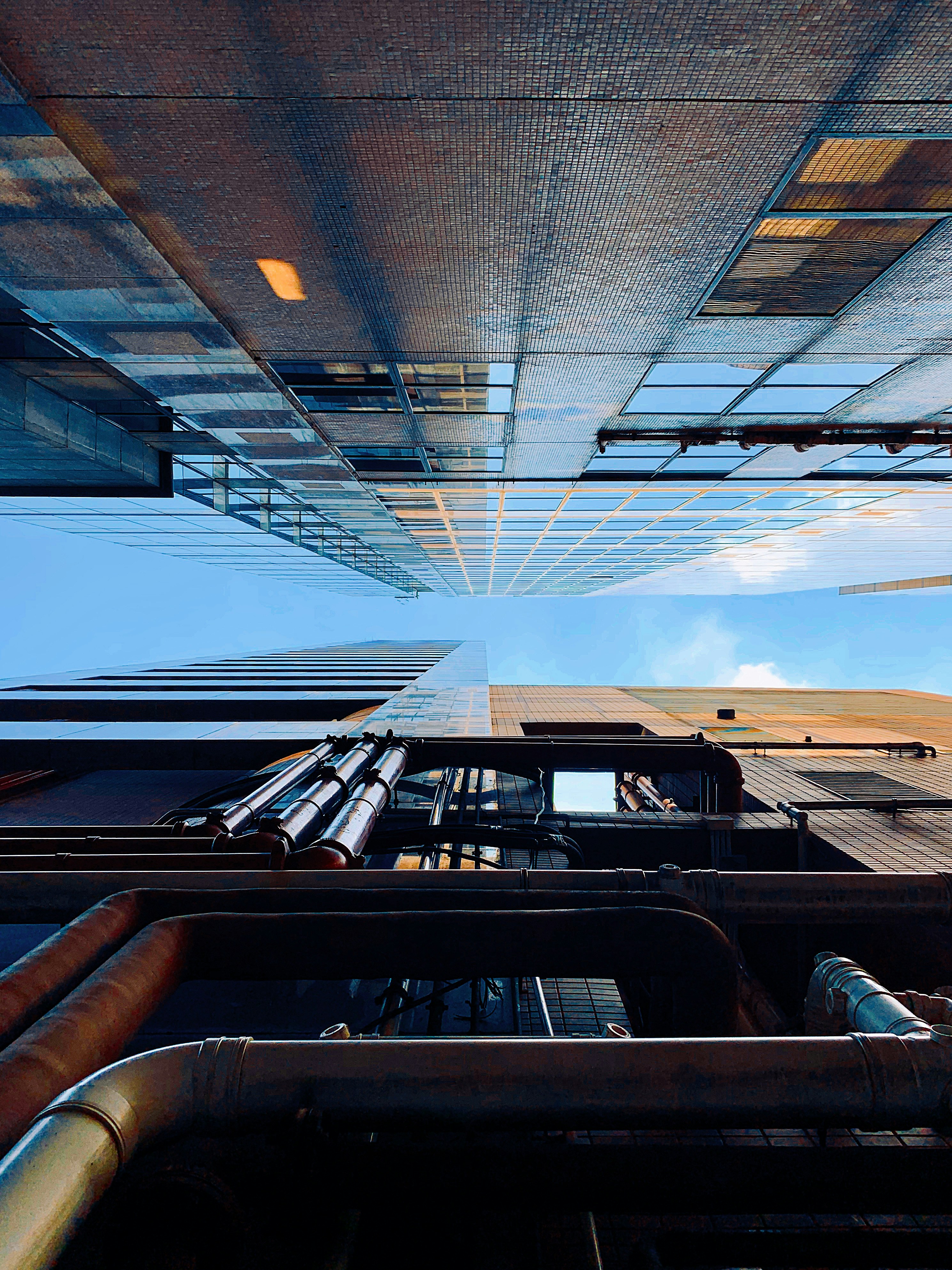 Looking up through a narrow alleyway, contrasting glass and brick buildings frame the bright sky above. Pipes and structural elements add depth to the urban scene.