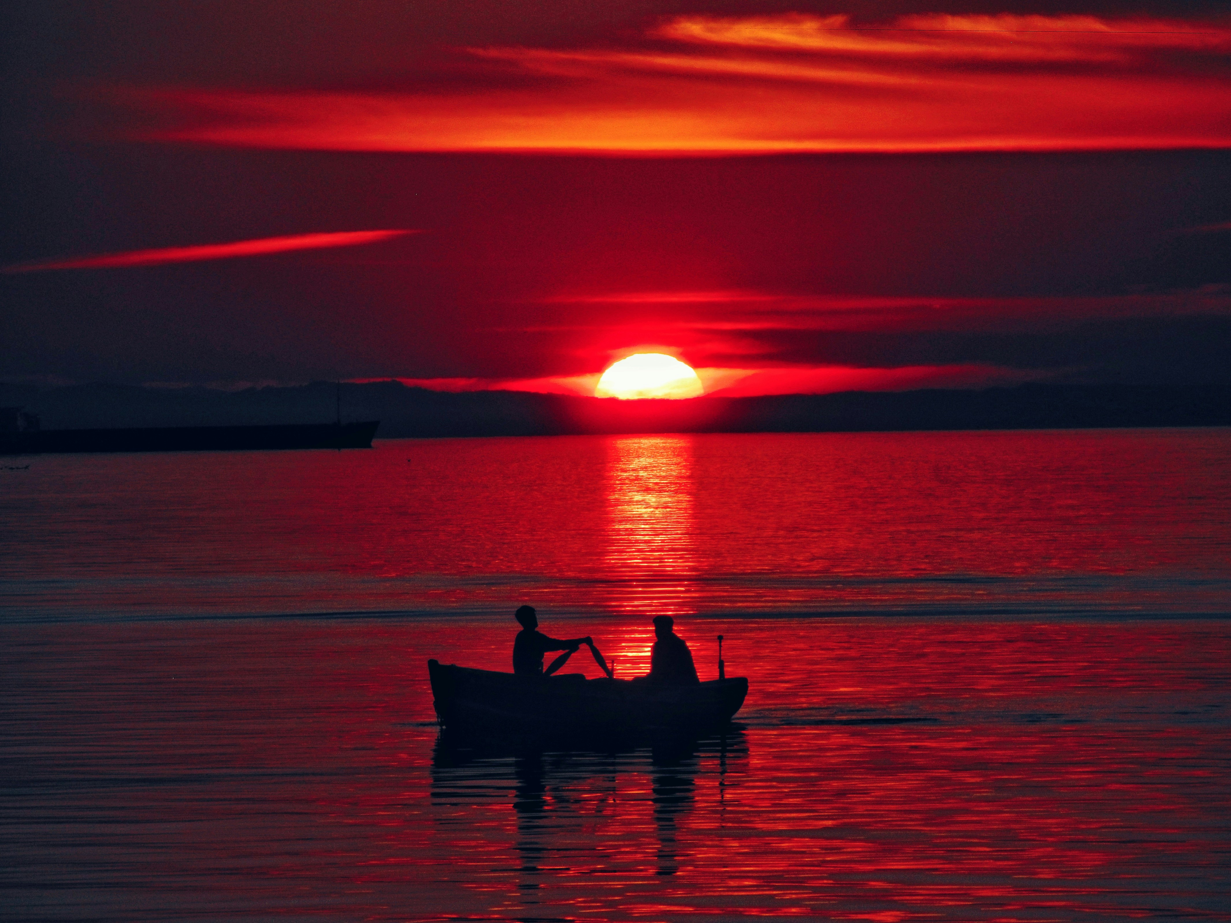 Two figures in a boat silhouetted against a vibrant sunset, with deep red and orange hues reflecting on the water's surface.