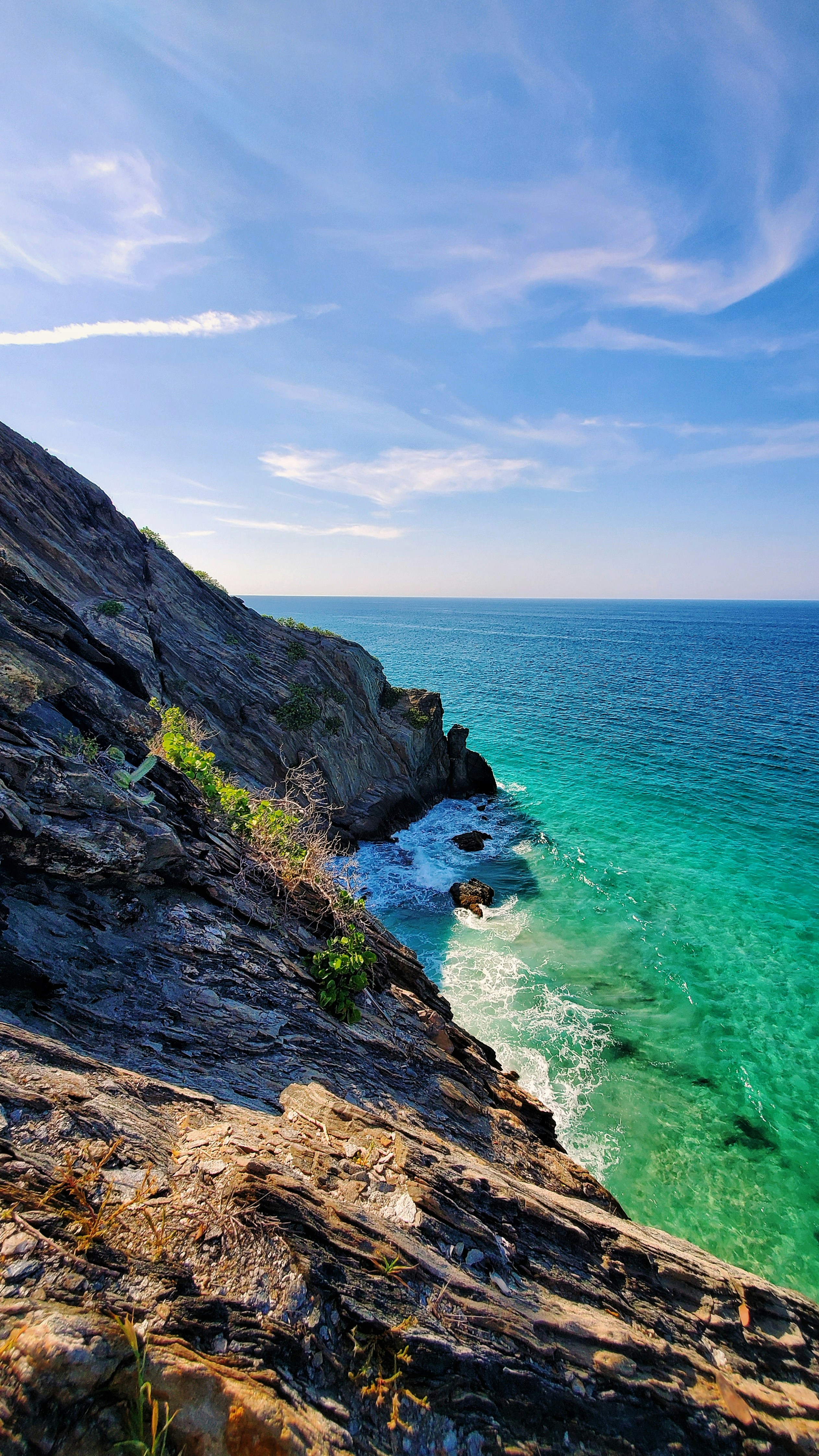 Vibrant turquoise waters crash against rocky cliffs under a clear blue sky. The lush greenery adds a touch of life to the rugged landscape.