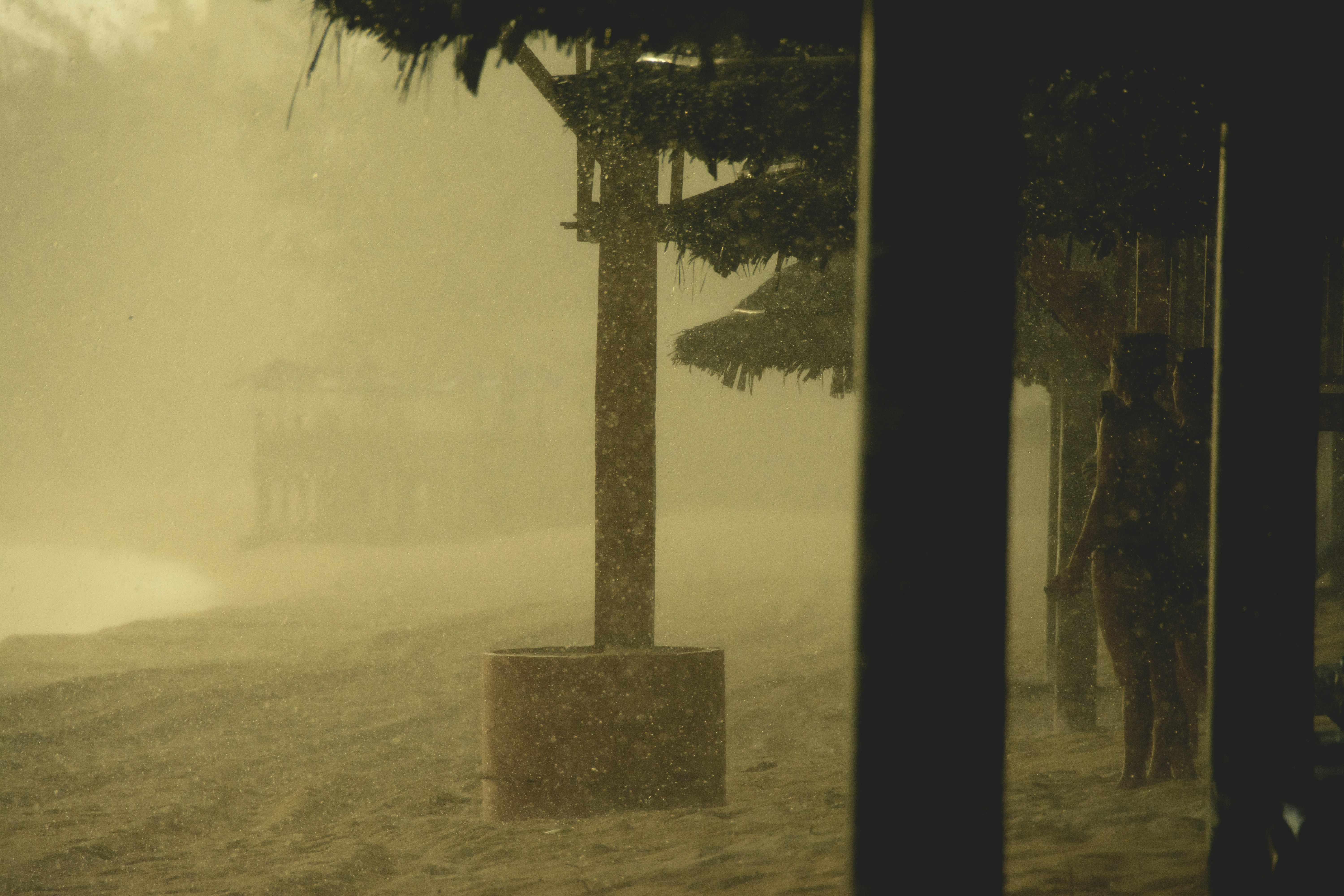 Raindrops cascade over a serene beach scene, framed by silhouetted palm structures and distant figures seeking shelter. The atmosphere is thick with mist and mystery.