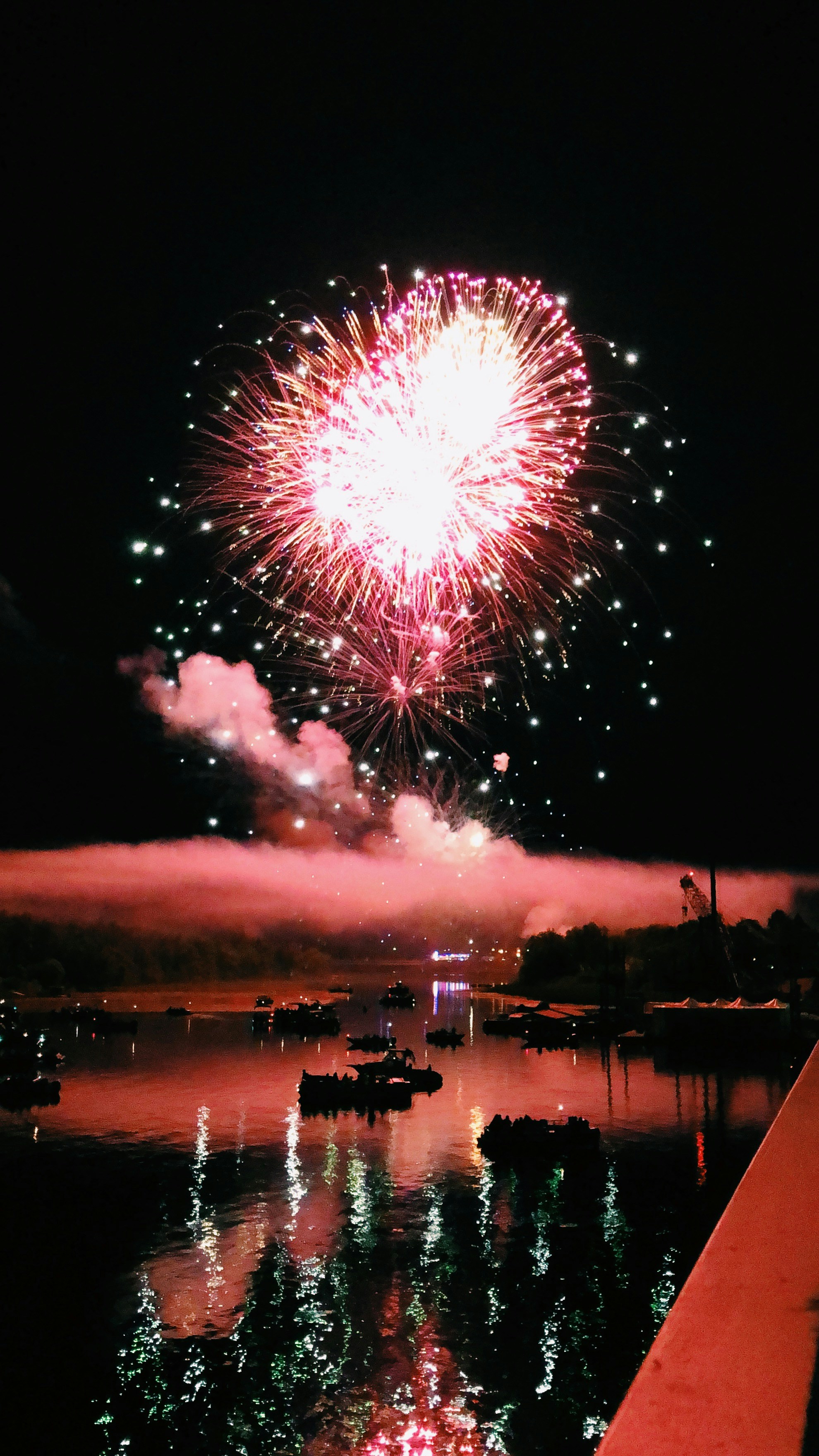 Vibrant fireworks display bursting in heart shapes over a tranquil river, reflecting the colorful lights and smoke in the water below.