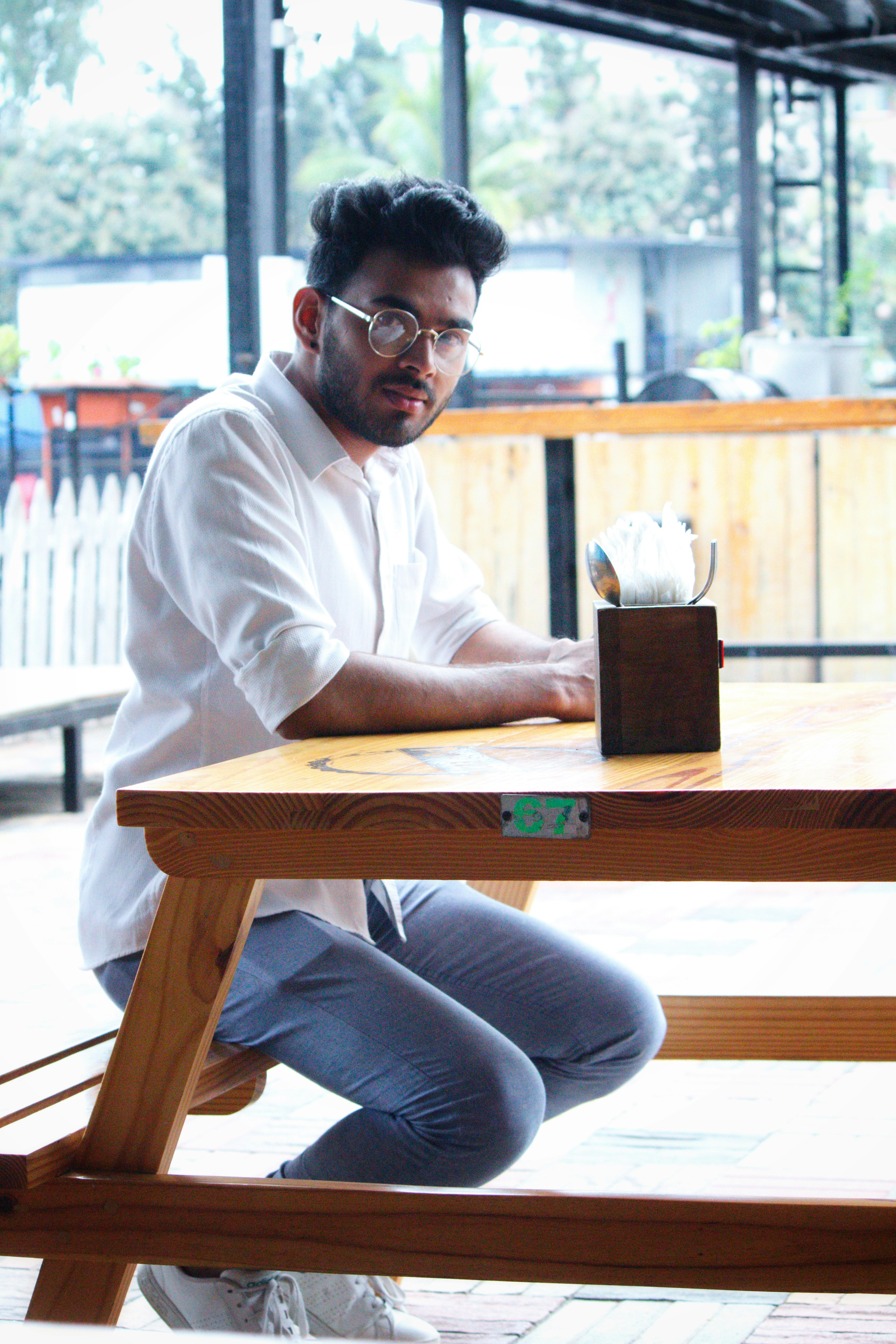 Young man seated at a wooden table in a café, with a unique holder for utensils in front of him. Natural light filters through the space, creating a relaxed atmosphere.
