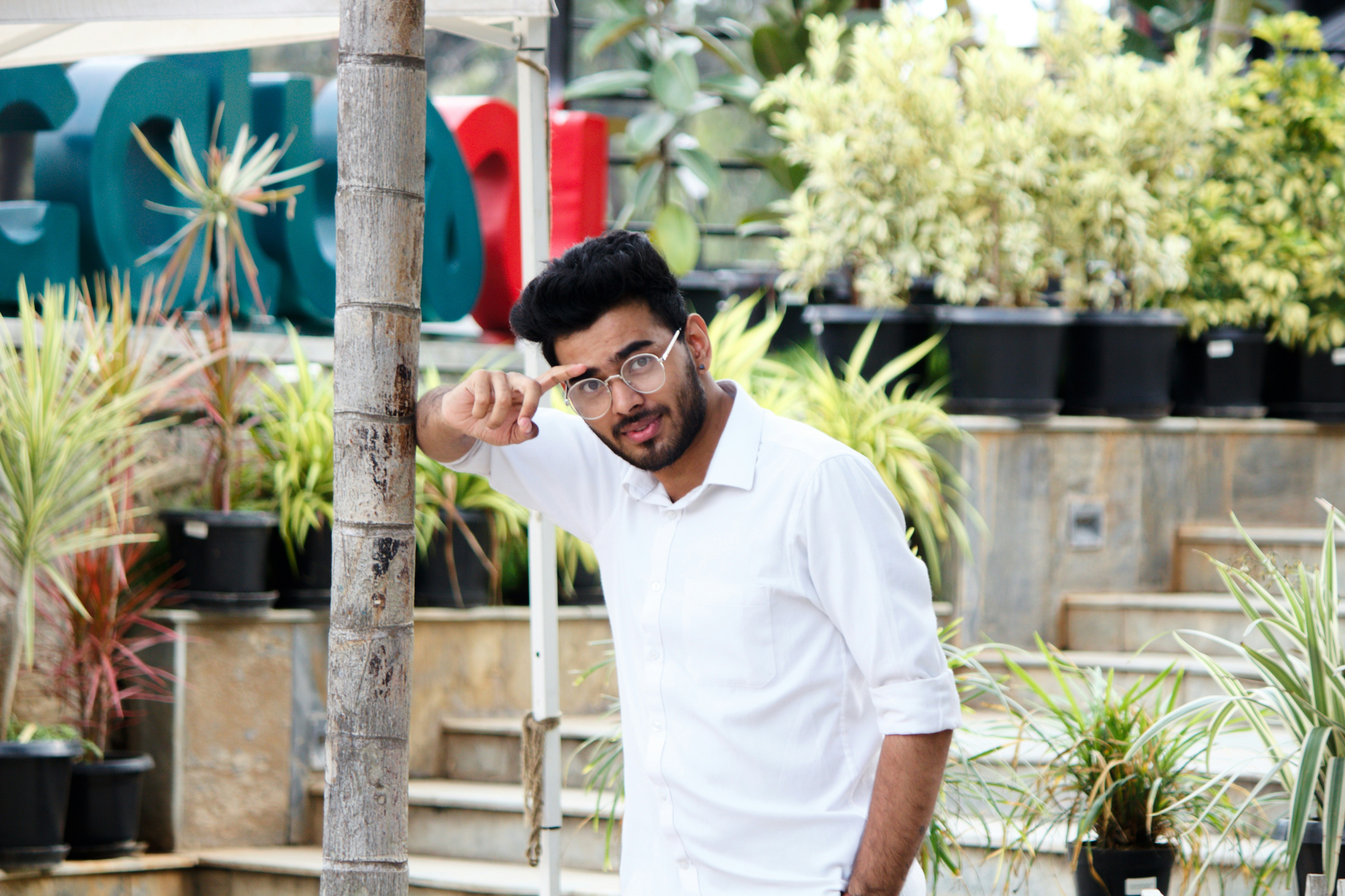 A young man poses thoughtfully in a garden filled with vibrant plants and decorative elements. His casual attire and relaxed demeanor add to the serene atmosphere.