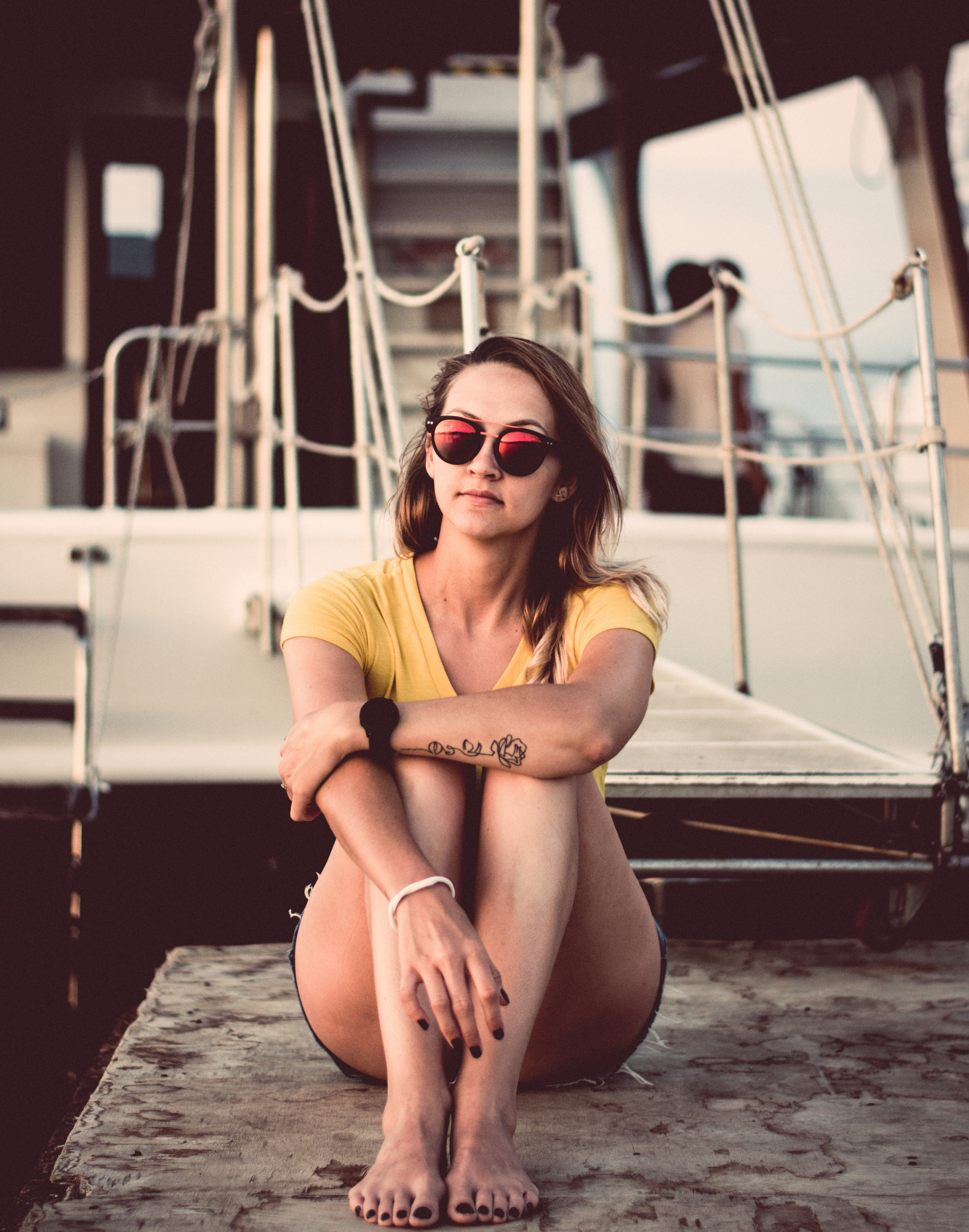 Young woman in yellow shirt and sunglasses sitting on a dock, reflecting in a tranquil setting with a boat in the background.