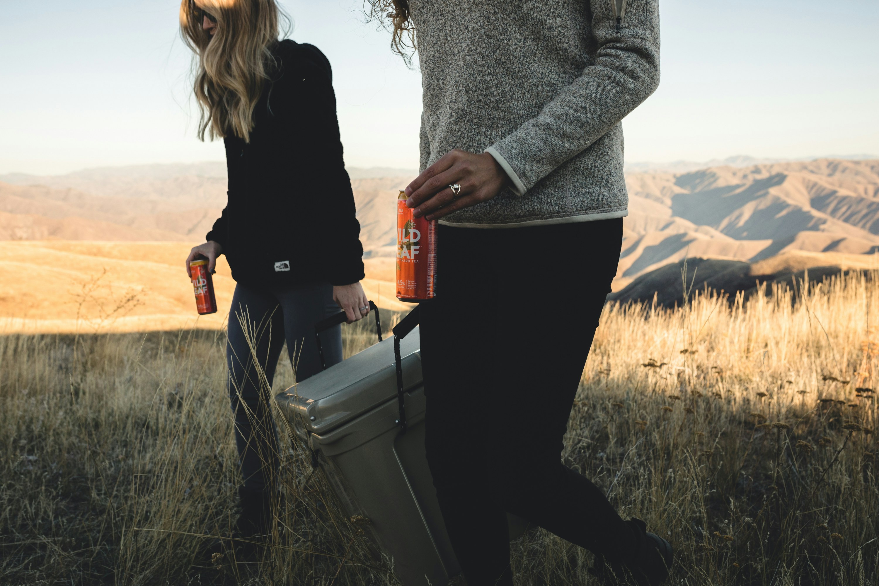 Two individuals trek across a golden hillside, each holding a beverage, embodying the spirit of outdoor exploration.