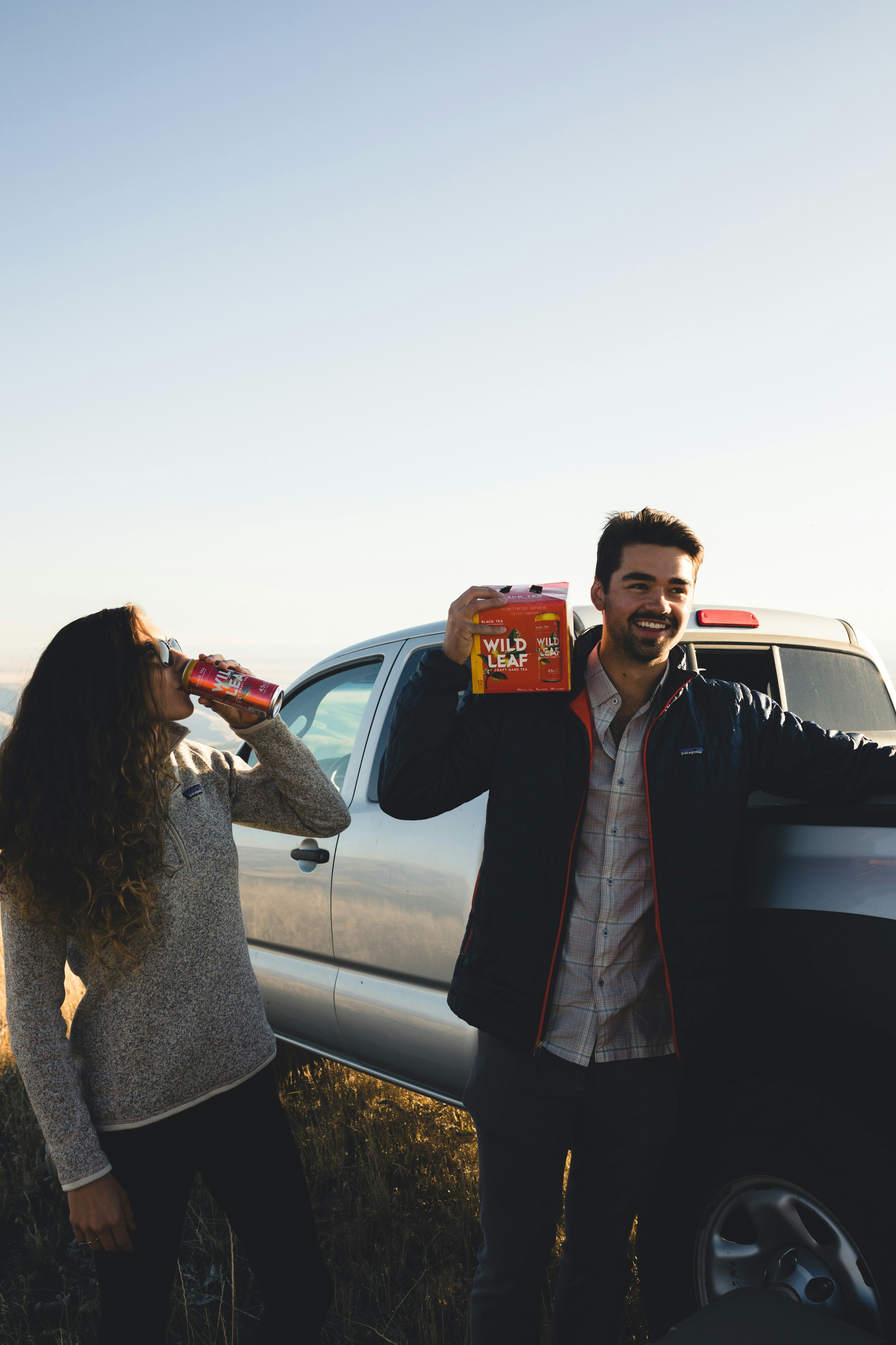 Two friends enjoying beverages outdoors beside a vehicle during sunset, embodying a spirit of camaraderie and adventure.