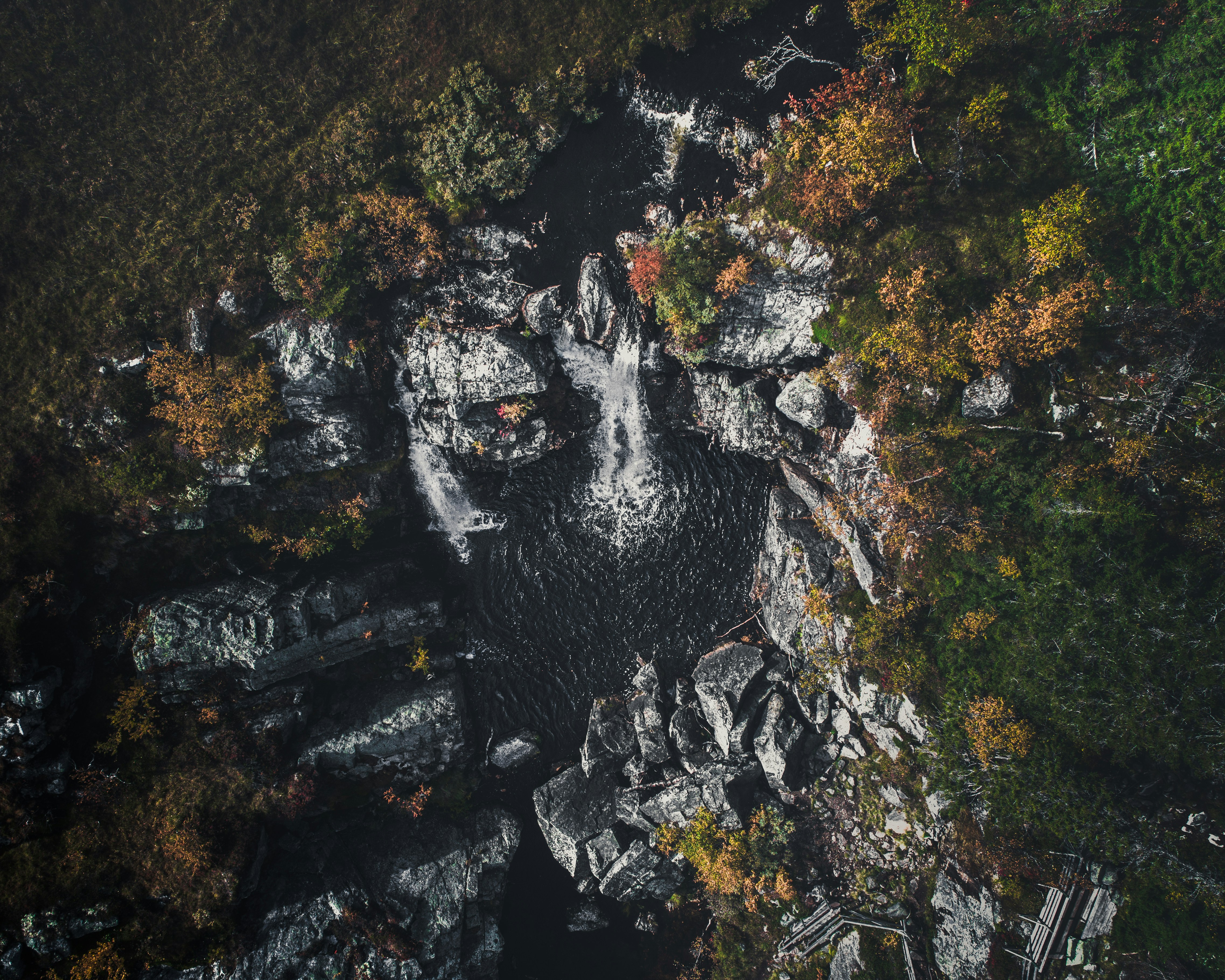 Aerial view of a rocky stream surrounded by autumn trees; water weaves through boulders creating a winding, high-contrast landscape.