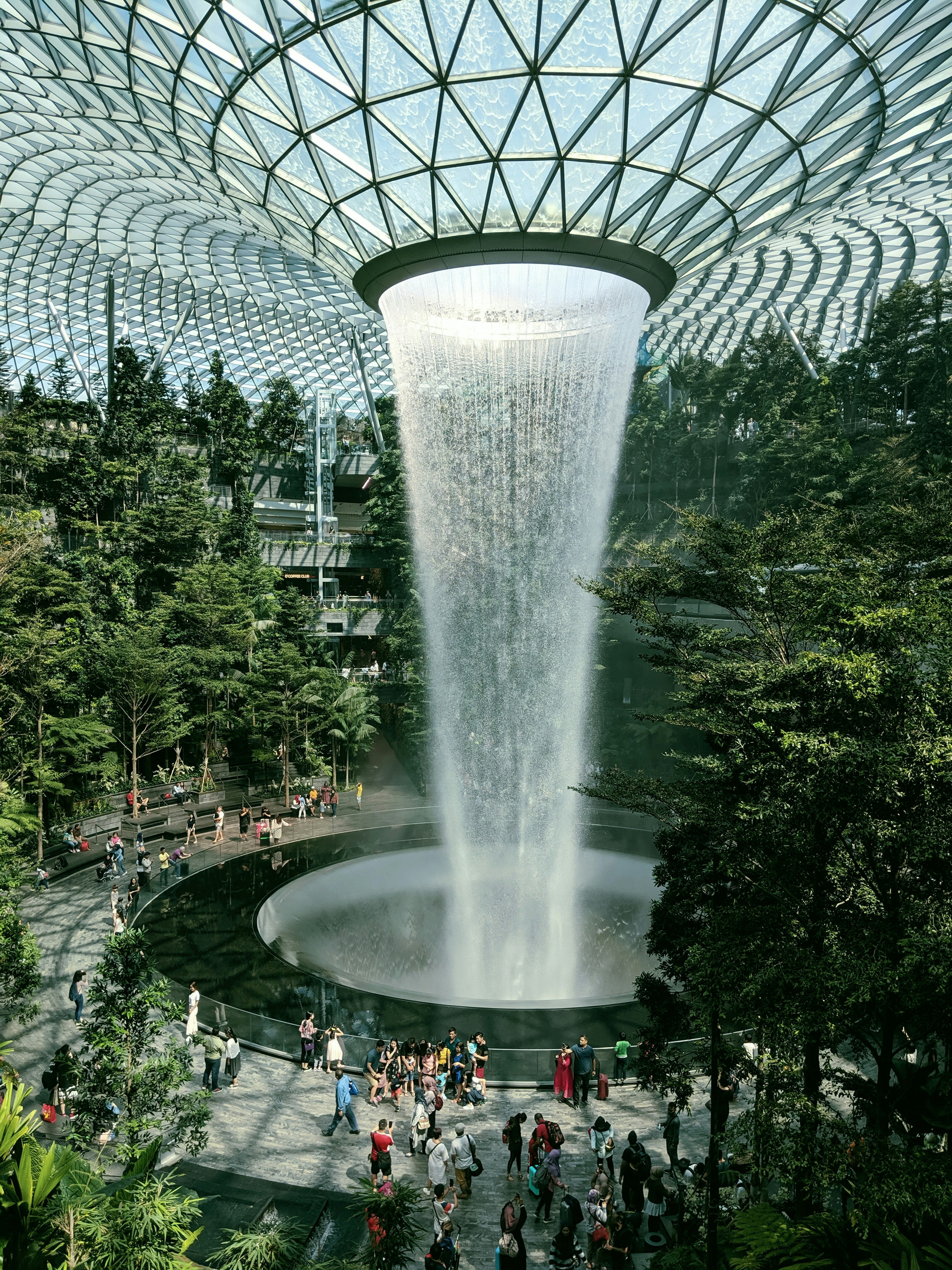A stunning indoor waterfall descends from a glass dome, surrounded by lush greenery and captivated visitors. The design harmoniously blends nature and architecture.