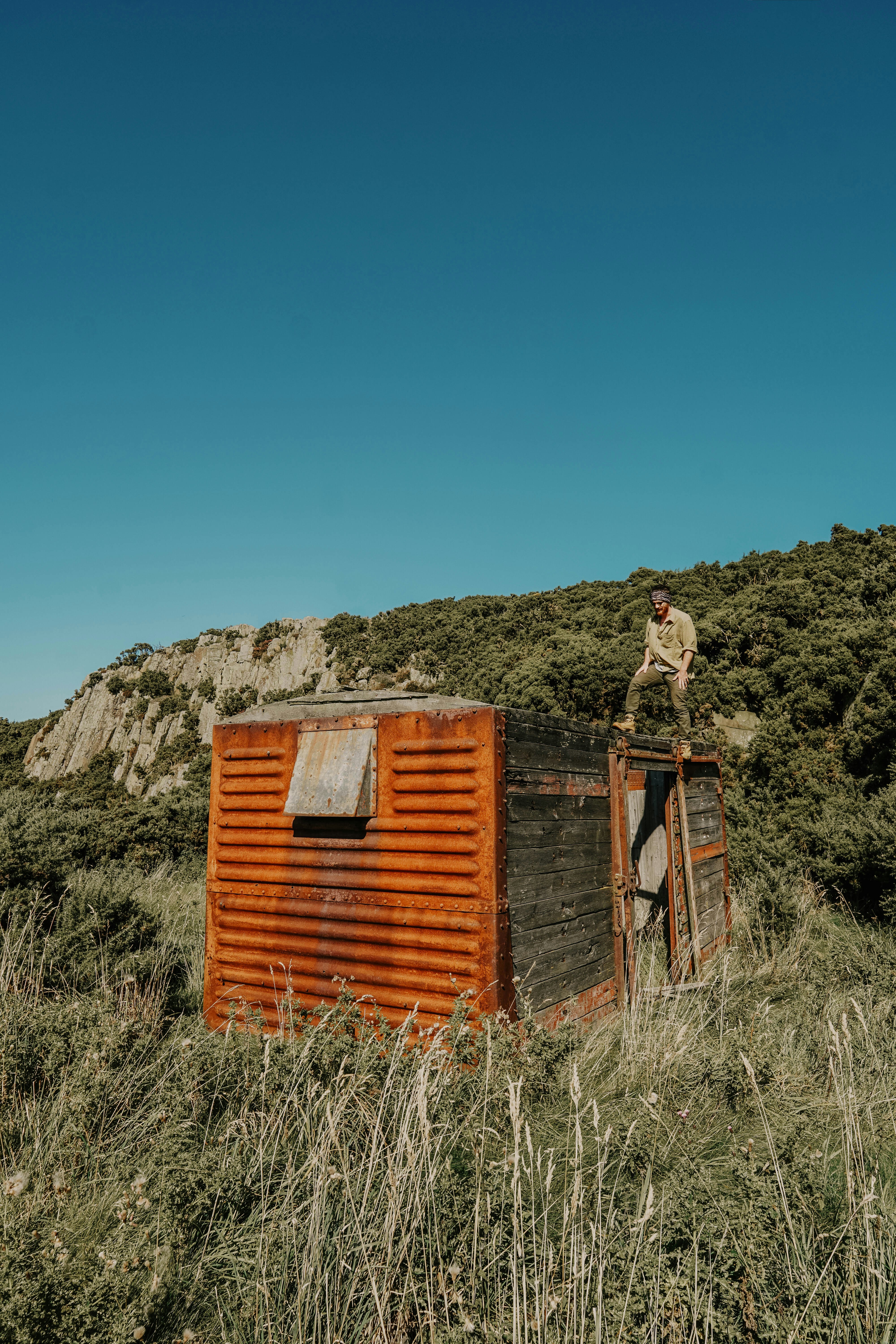 A person stands atop a weathered structure, surrounded by lush greenery and rocky hills under a clear blue sky.