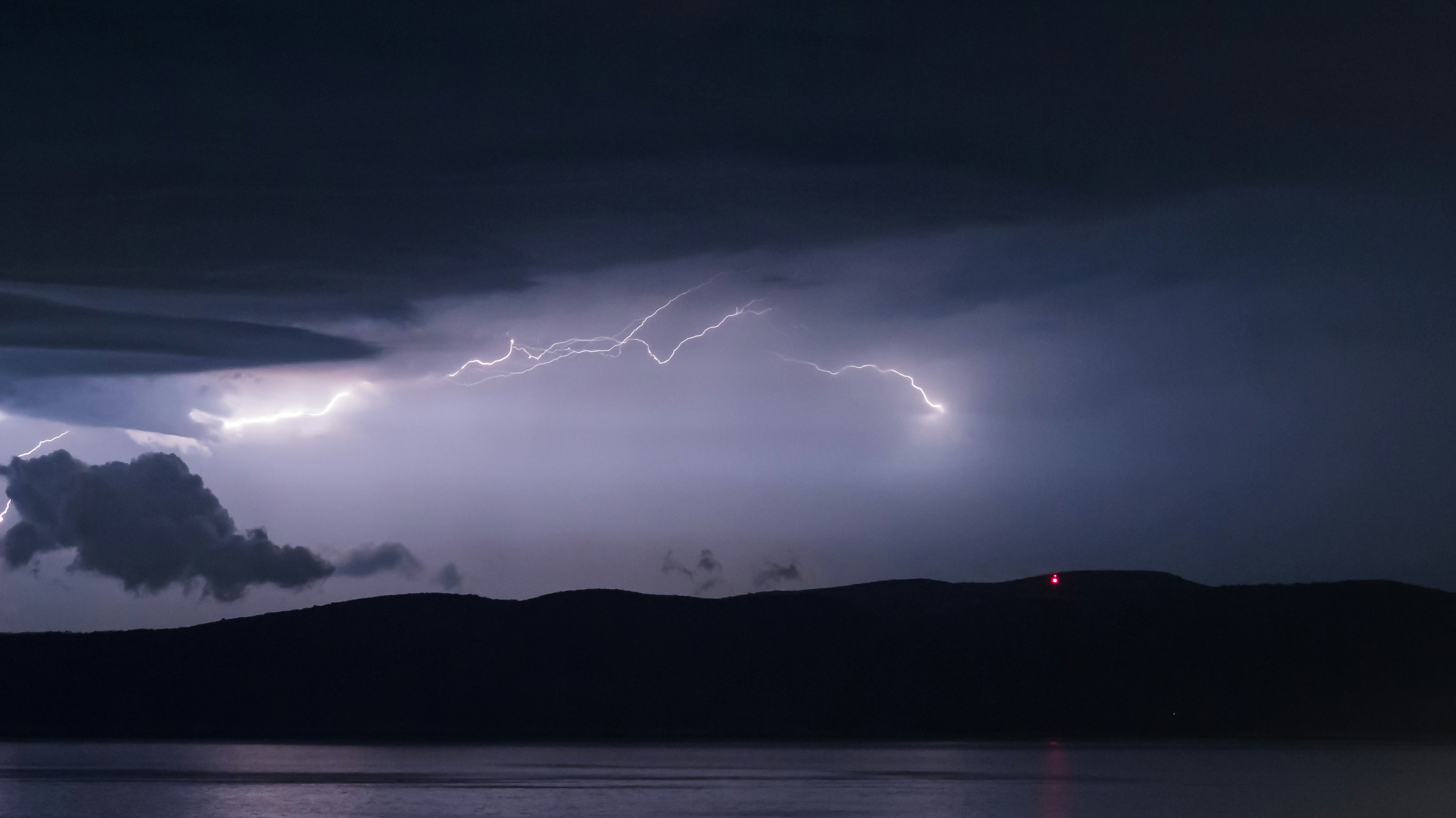 Dramatic lightning strikes illuminate a stormy sky over a tranquil lake, with distant mountains silhouetted against the vibrant backdrop.