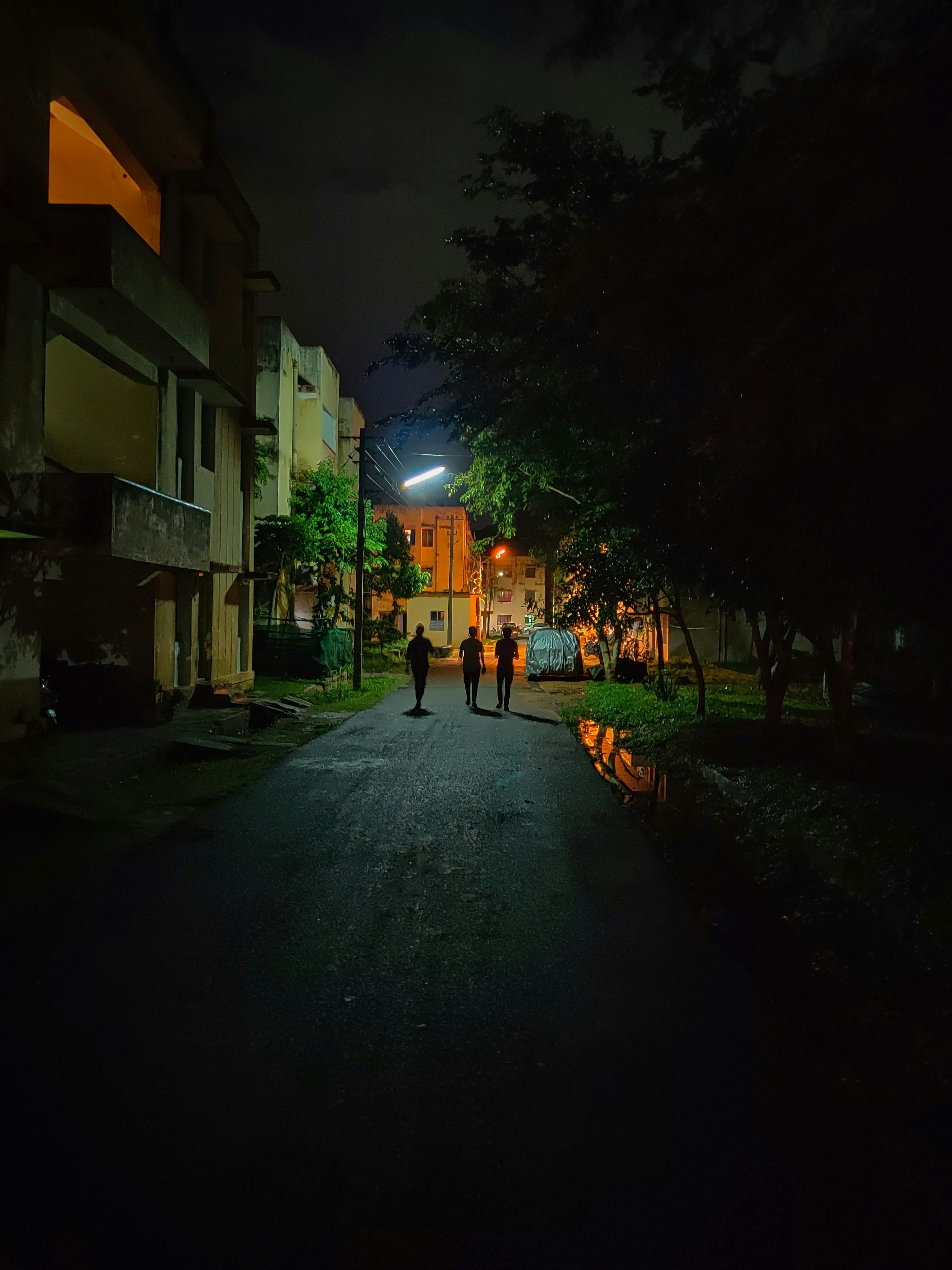 Three figures walking down a dimly lit street flanked by buildings and trees at night. Streetlights cast a warm glow on the pavement.