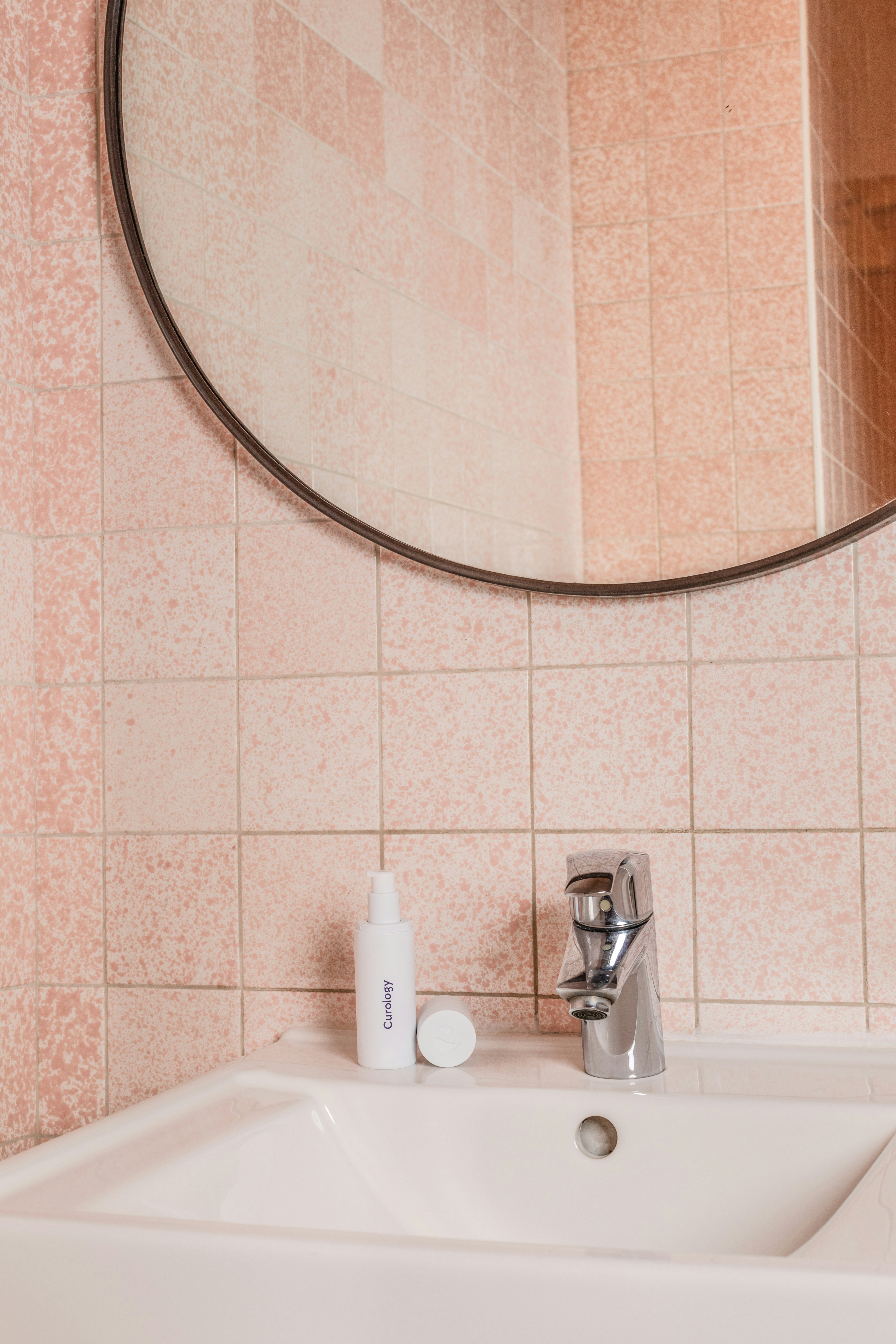 A modern bathroom sink with a sleek faucet and skincare products on the counter, framed by a round mirror against a soft pink tiled wall.