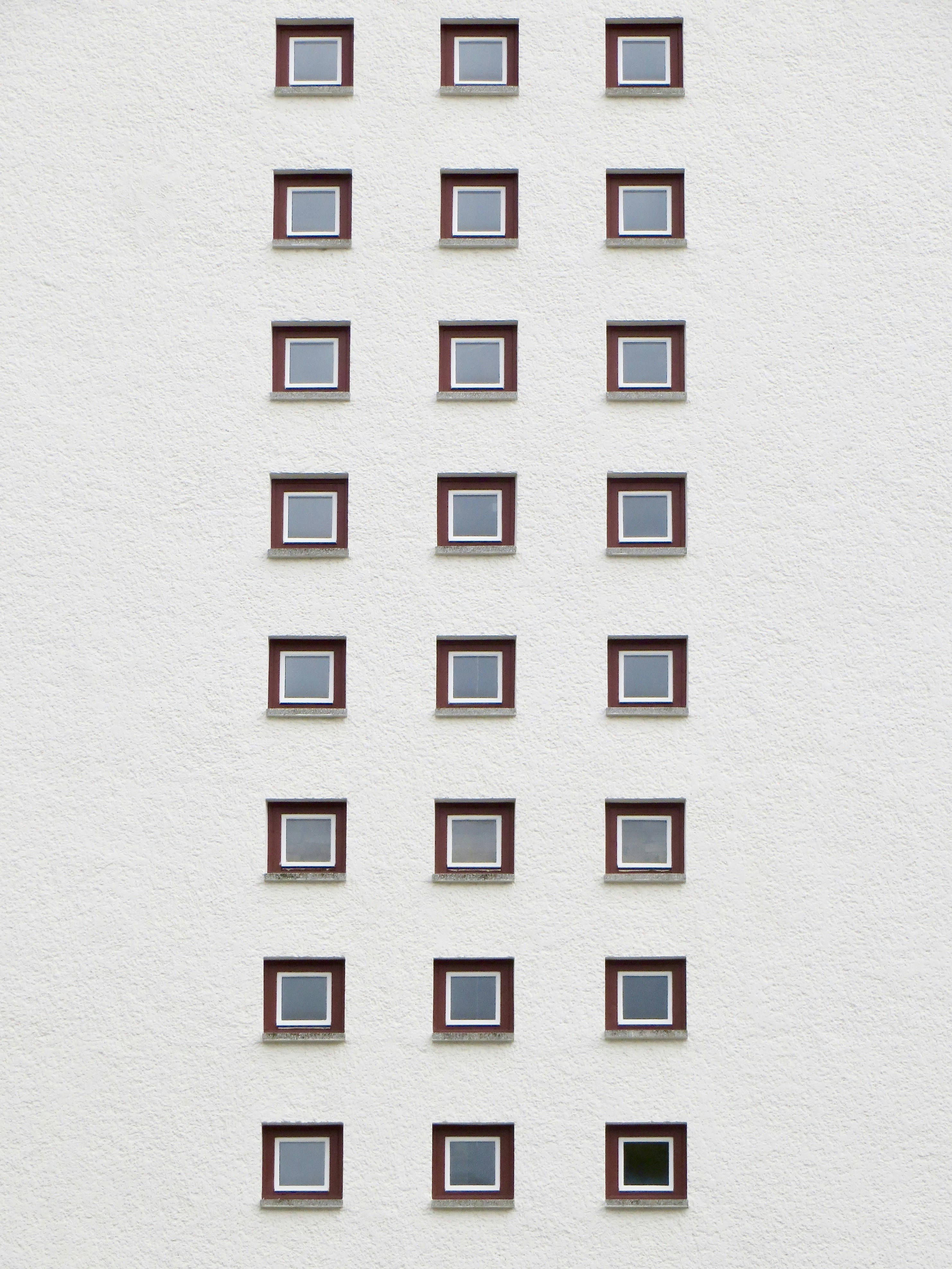 Photograph of a pale textured wall featuring a strict grid of burgundy-framed square windows.