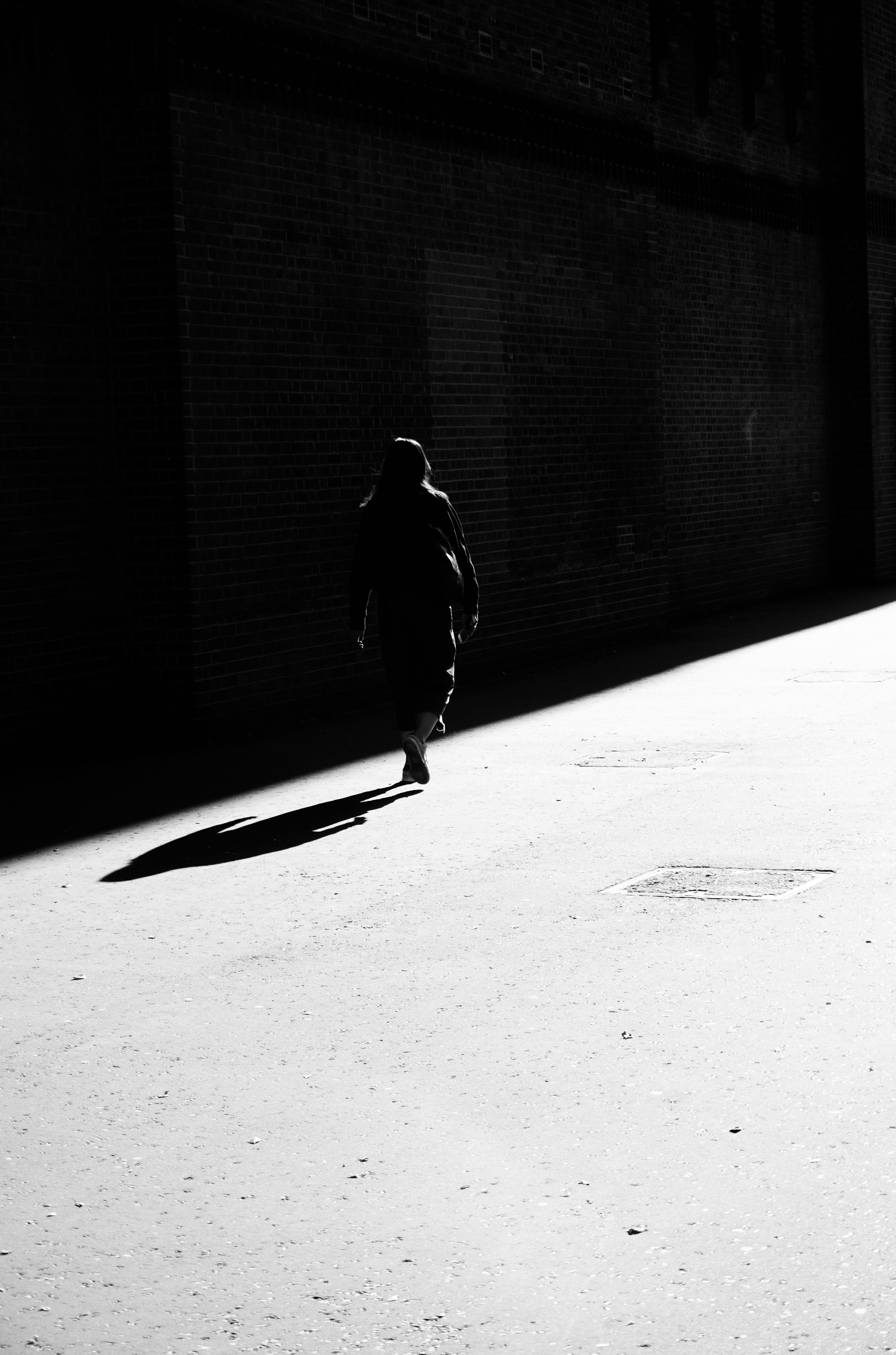 A figure walks alone in a dimly lit urban space, casting a long shadow on the ground. The stark contrast of light and dark creates a dramatic atmosphere.