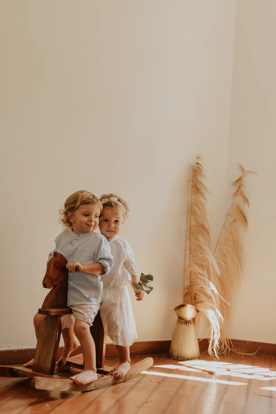 Two young children sit close on a wooden rocking horse against a sunlit wall, one holding a small bouquet; warm, natural-hour tones and soft shadows define the scene.