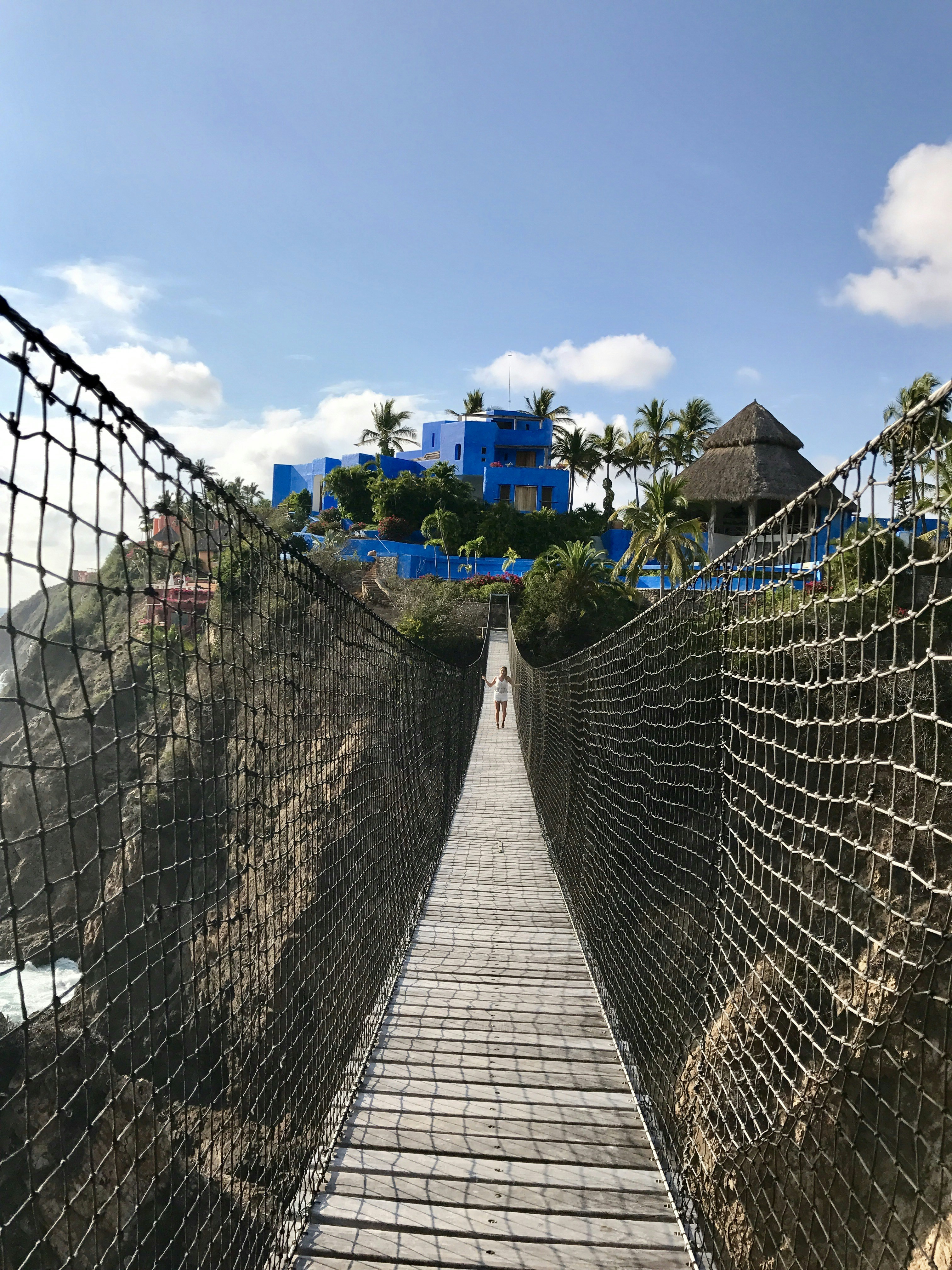 Suspended wooden bridge leading to a vibrant blue house surrounded by palms, set against a clear sky. The scene captures the essence of coastal tranquility.