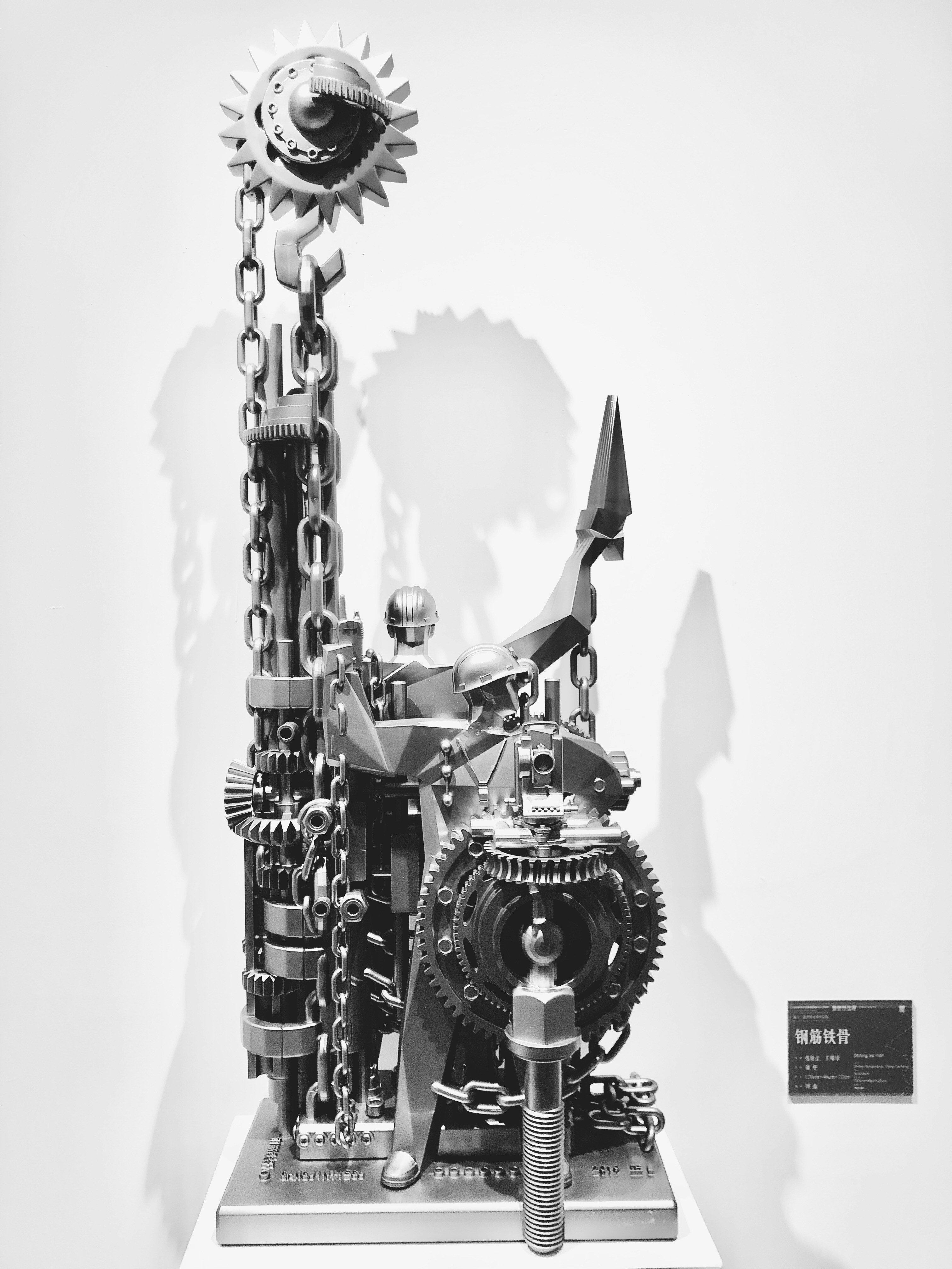 Intricate metallic sculpture featuring gears, chains, and a stylized figure, showcasing the blend of mechanical design and artistic expression.