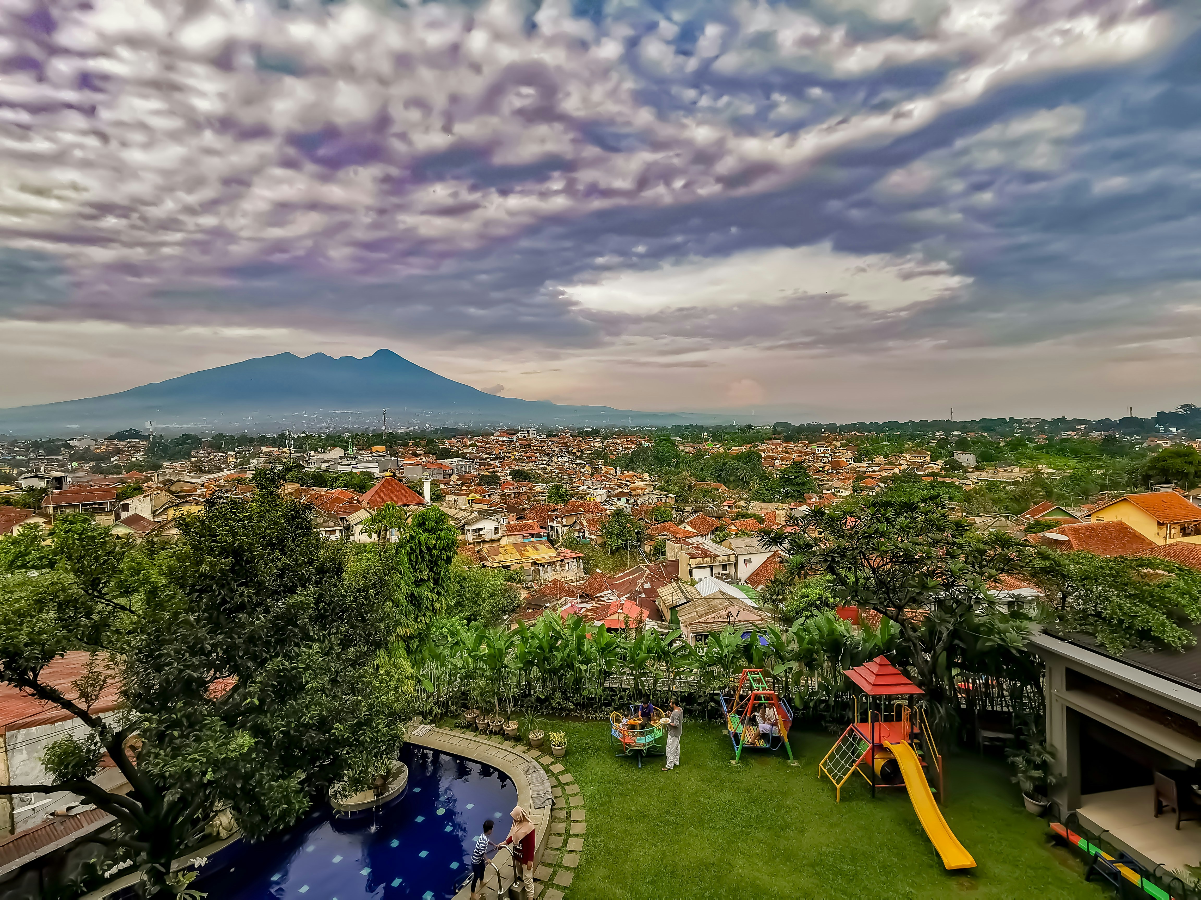 Expansive cityscape with Mt. Salak under a dramatic cloud-filled sky, bordered by greenery and a playground.