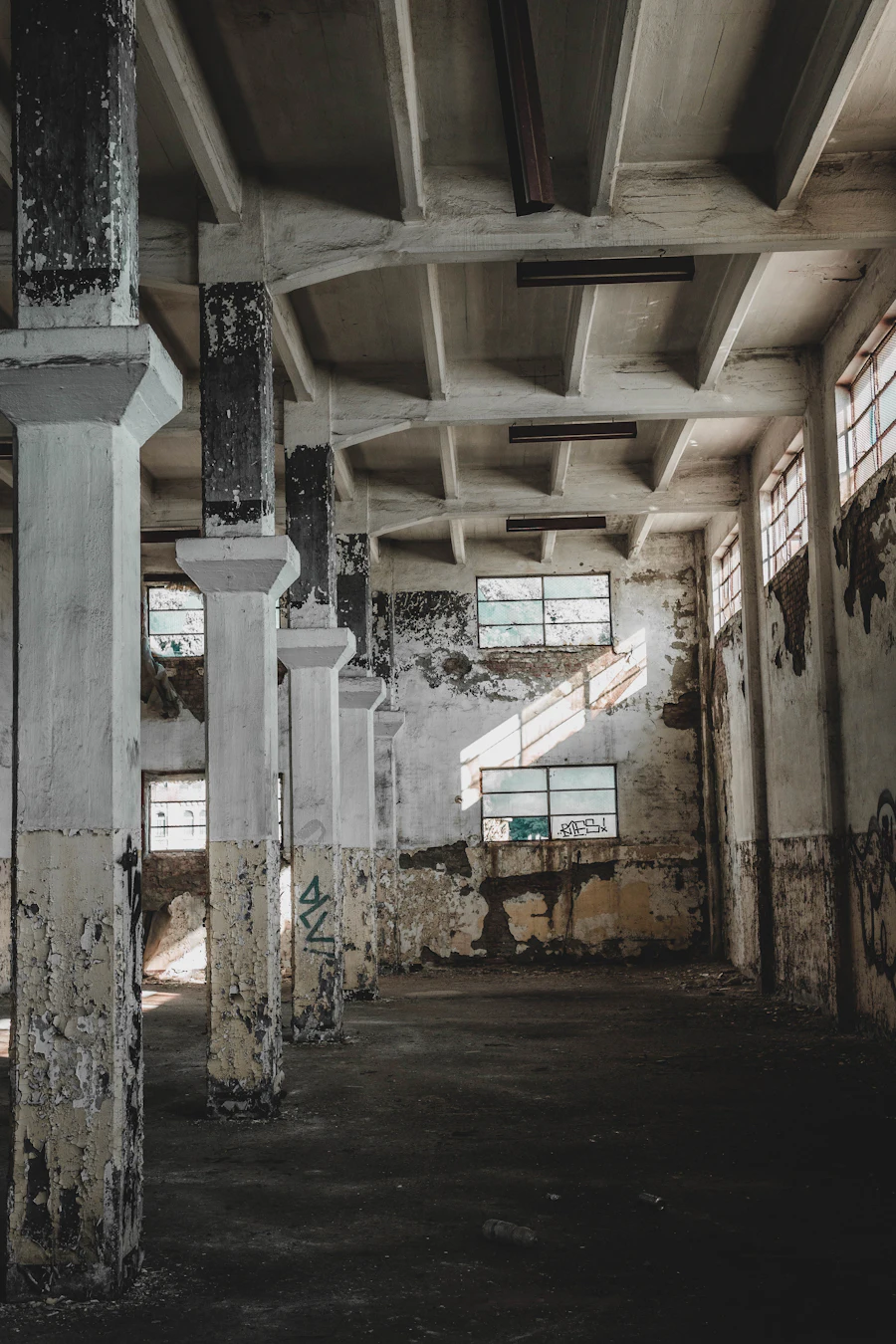 An abandoned factory interior bathed in angled sunlight that reveals peeling walls and weathered columns. The large windows cast geometric light patterns across a concrete floor, emphasizing texture and decay.