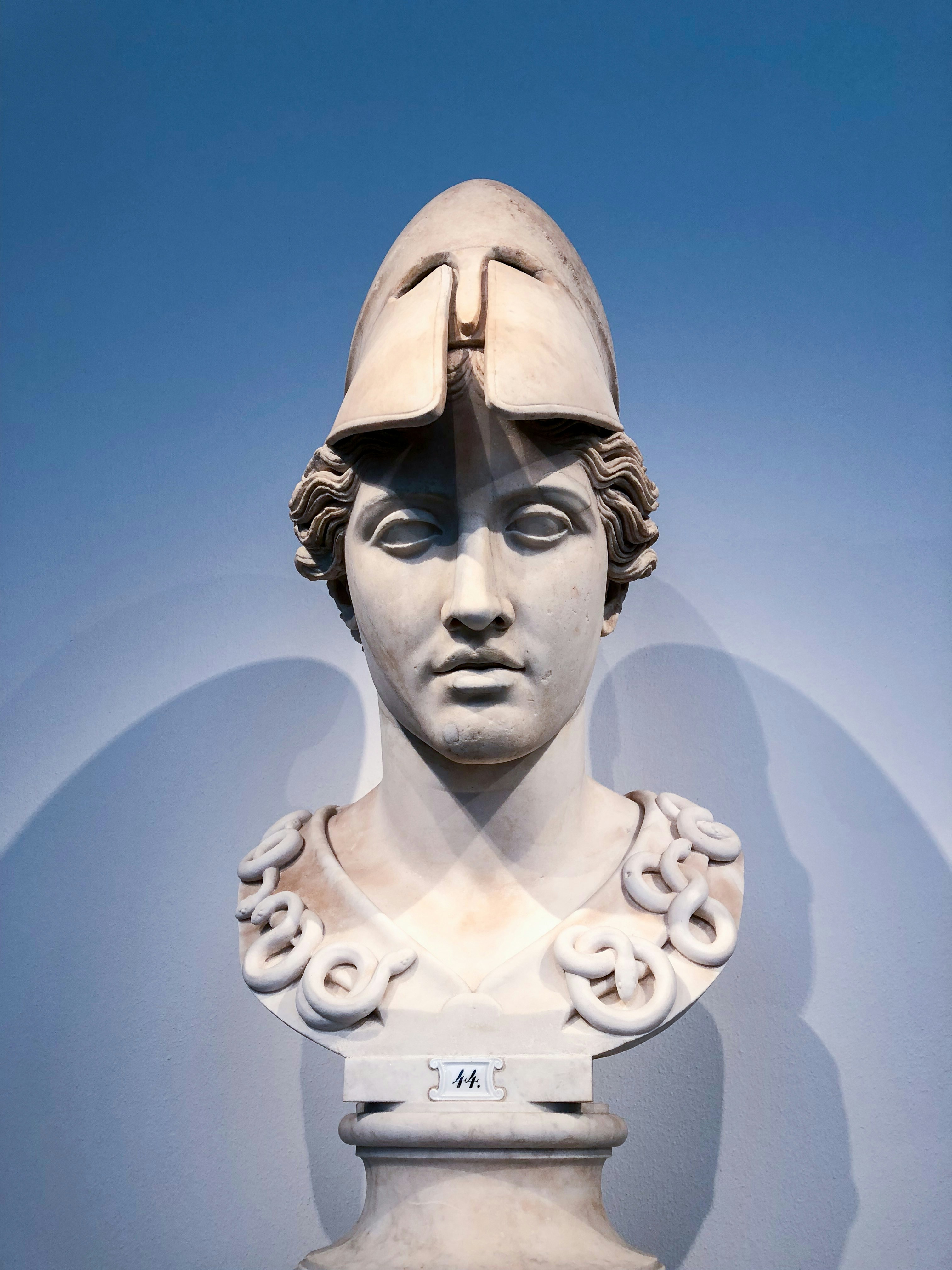 Marble bust of a helmeted figure adorned with serpentine details against a soft blue background.