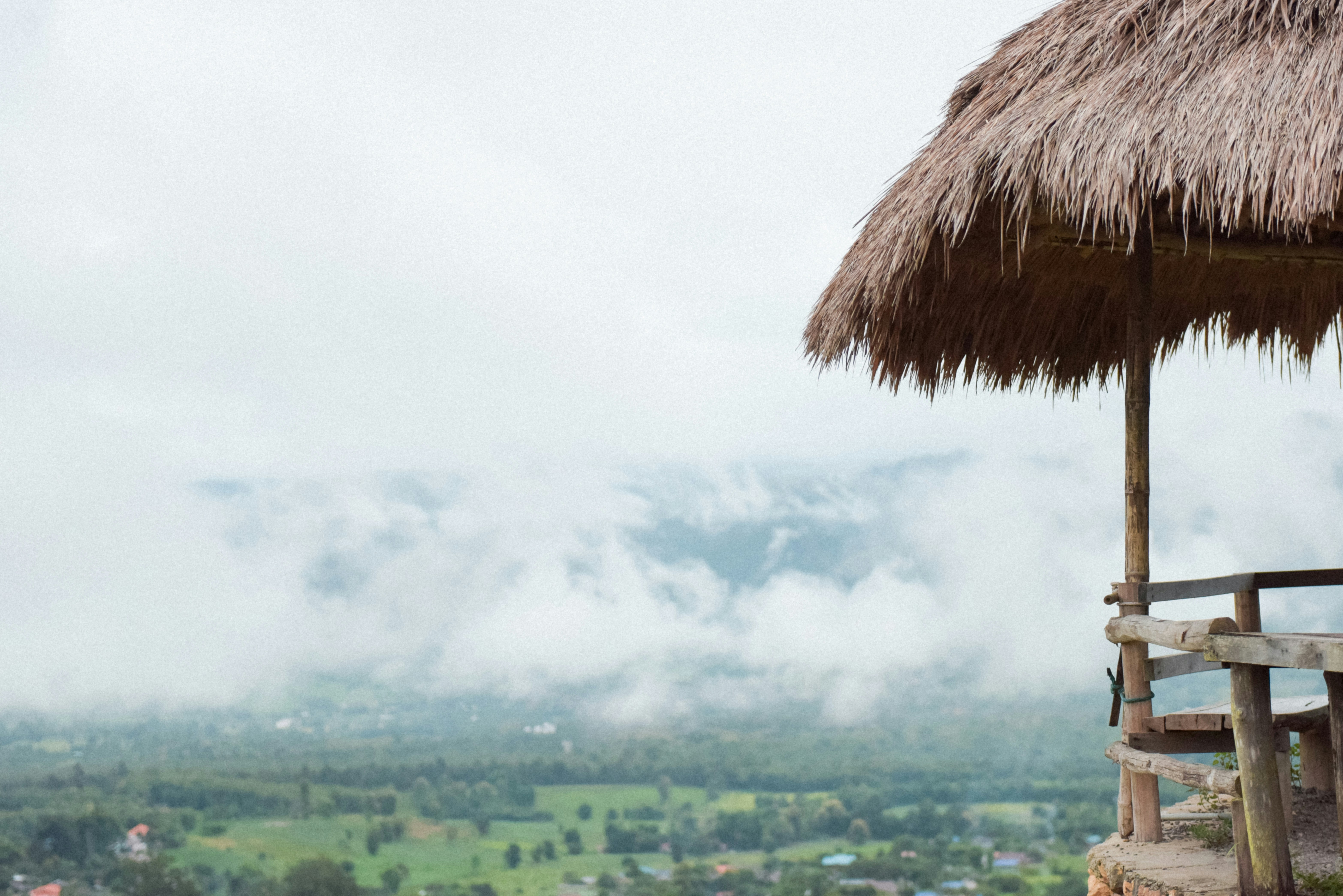 A rustic thatched gazebo perched on a hillside, overlooking a lush green valley shrouded in mist and clouds.