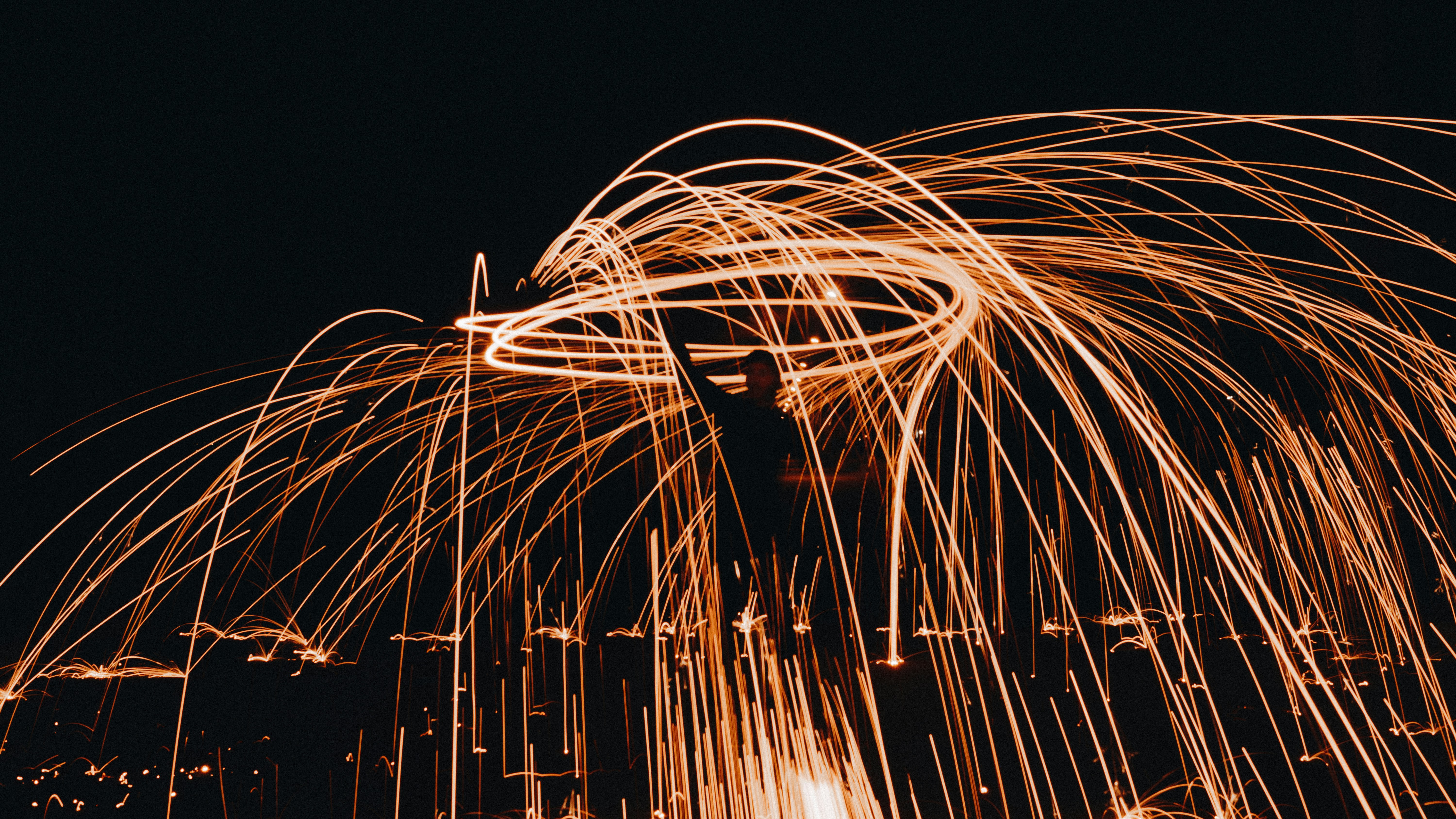 A figure spins a fiery steel wool display against a dark backdrop, creating a mesmerizing pattern of light trails.