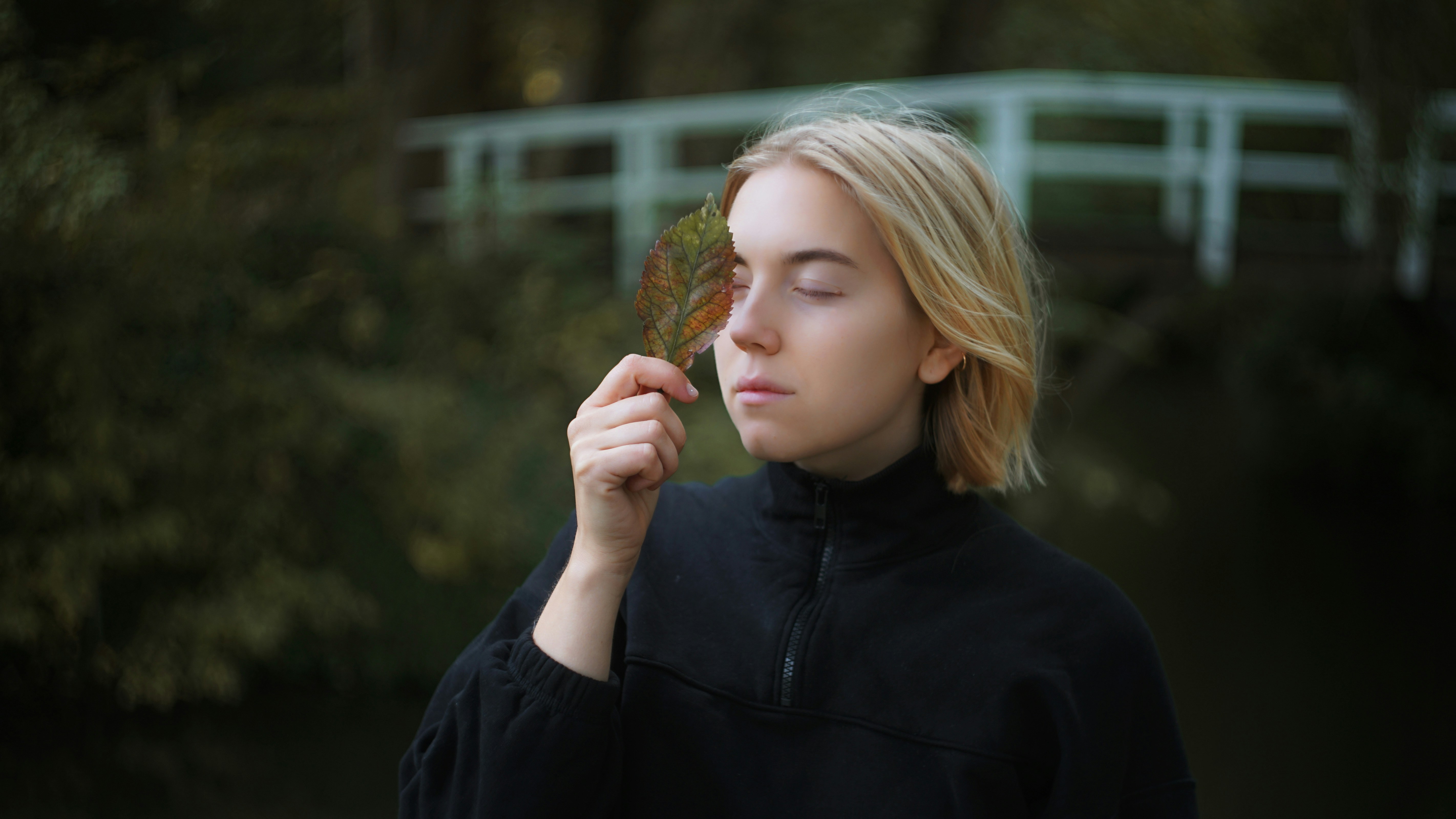 Young woman holding a leaf close to her face, eyes closed in contemplation, with a soft-focus background of greenery and a bridge.