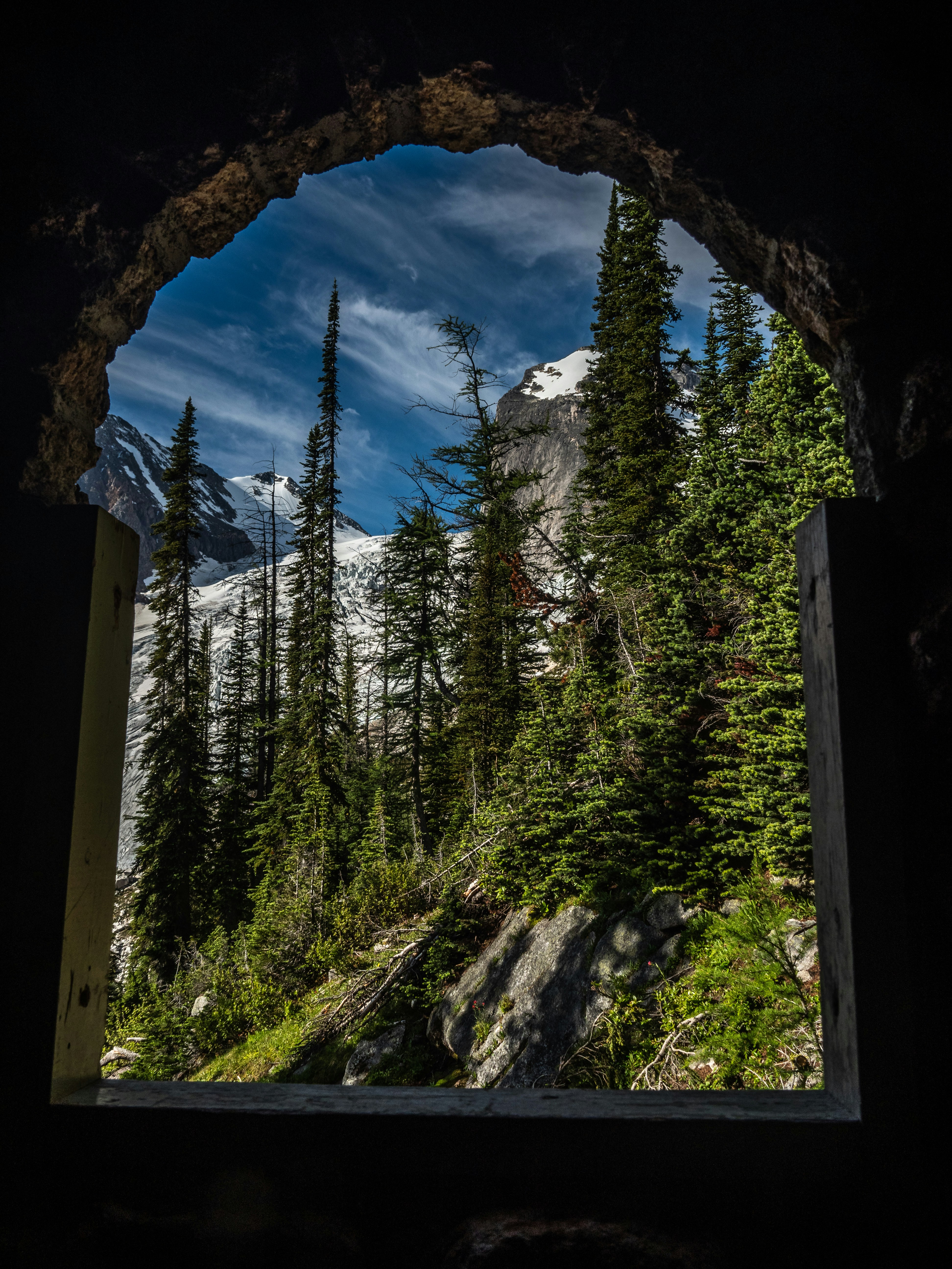 Snow-capped peaks framed by evergreen pines seen from a cave window