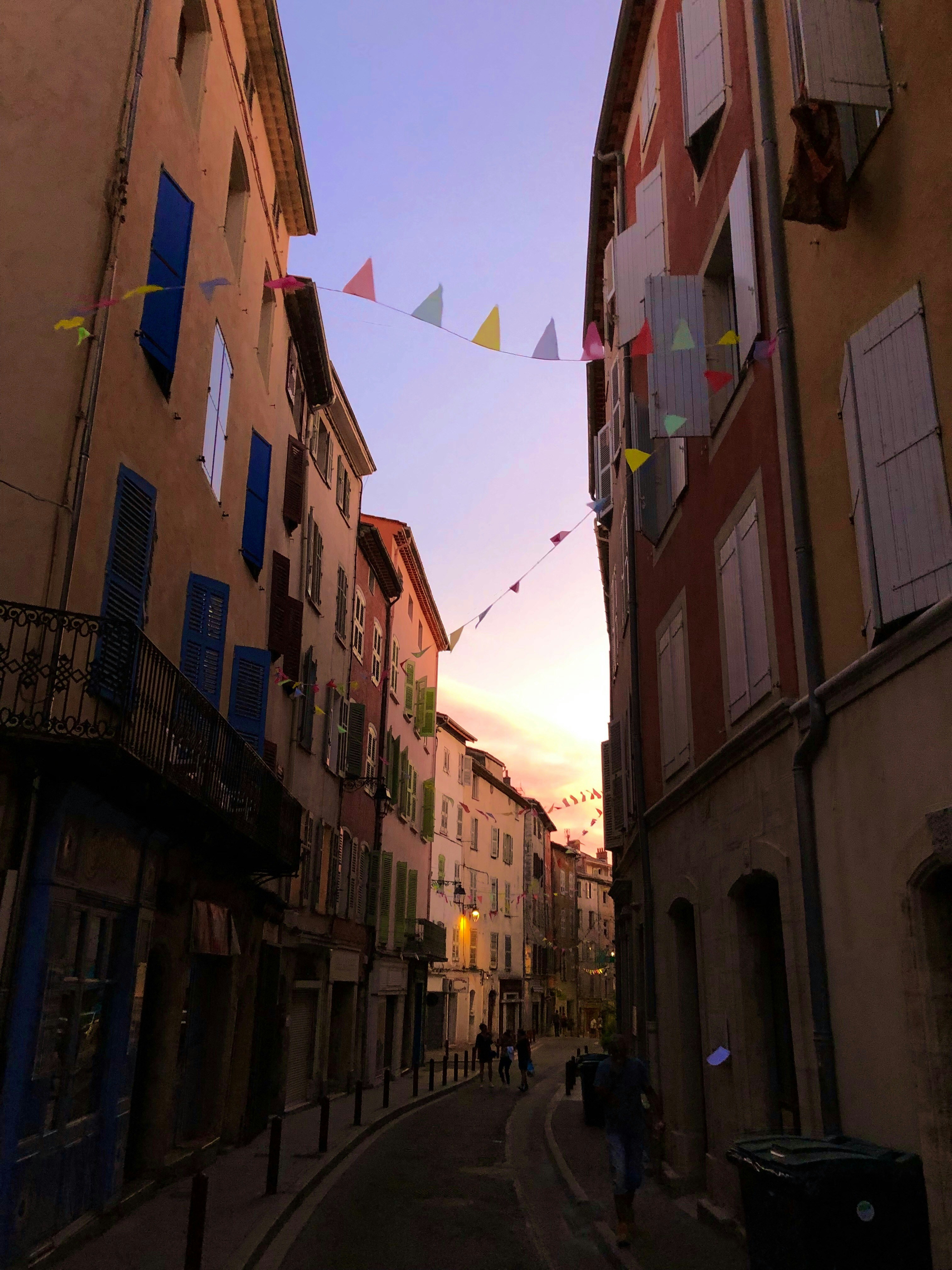 Charming narrow street in a French town adorned with colorful bunting under a twilight sky. Warm light spills from a window, inviting evening strolls.