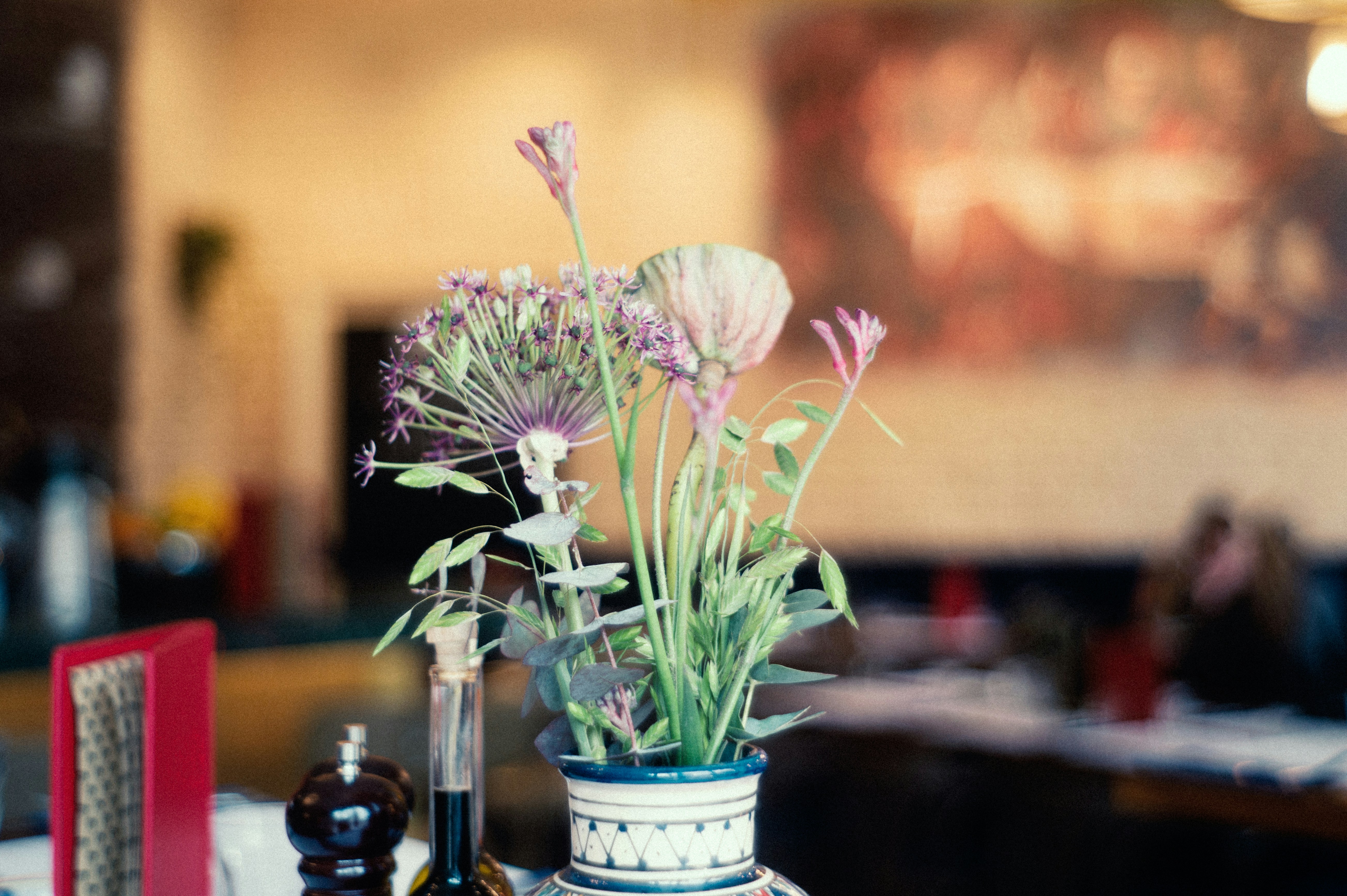 Bouquet of purple-pink flowers in a patterned ceramic vase sits on a coffee shop table with a warm, blurred background.