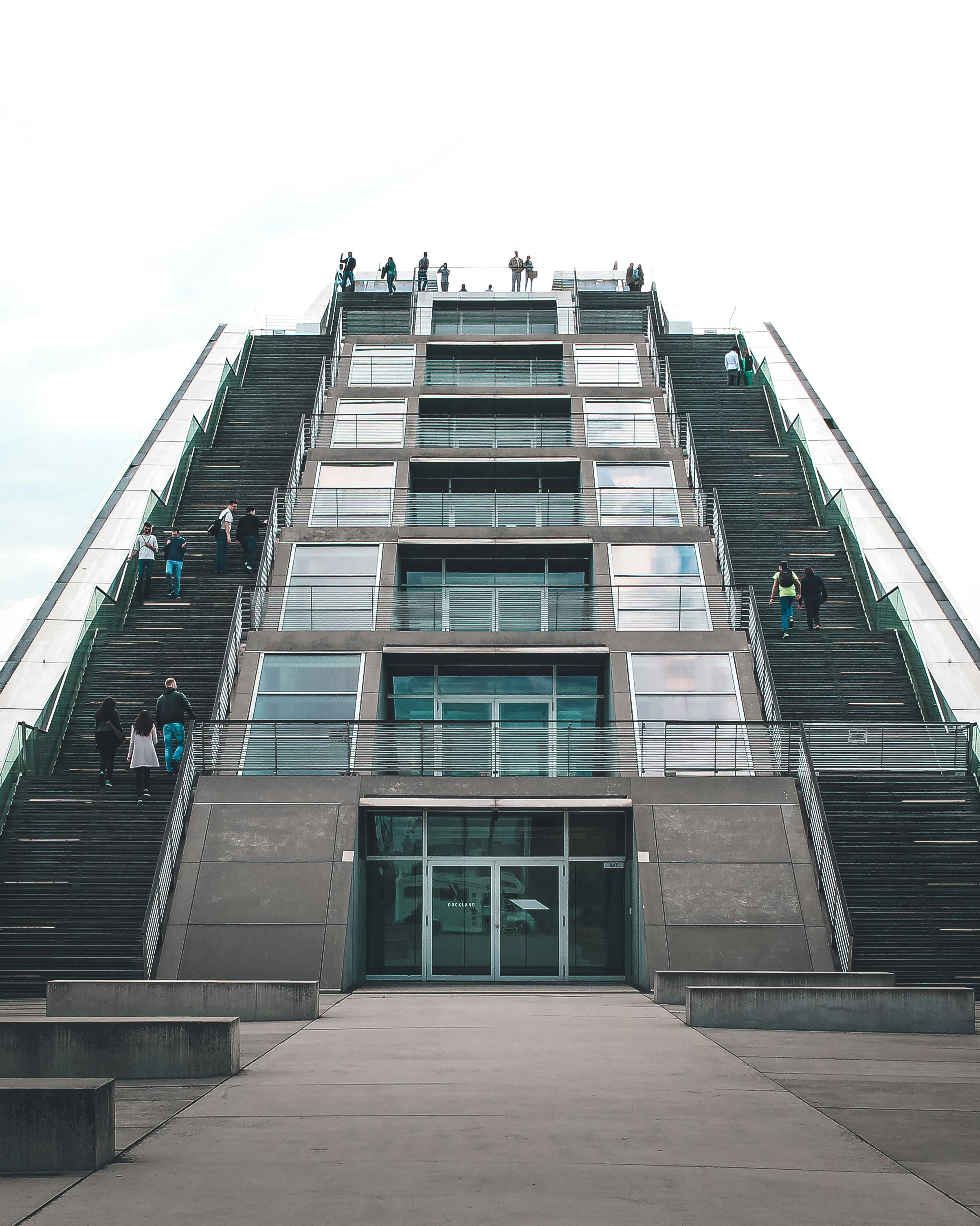 A striking modern staircase leads upward, flanked by glass walls and bustling visitors. The design emphasizes verticality and transparency.