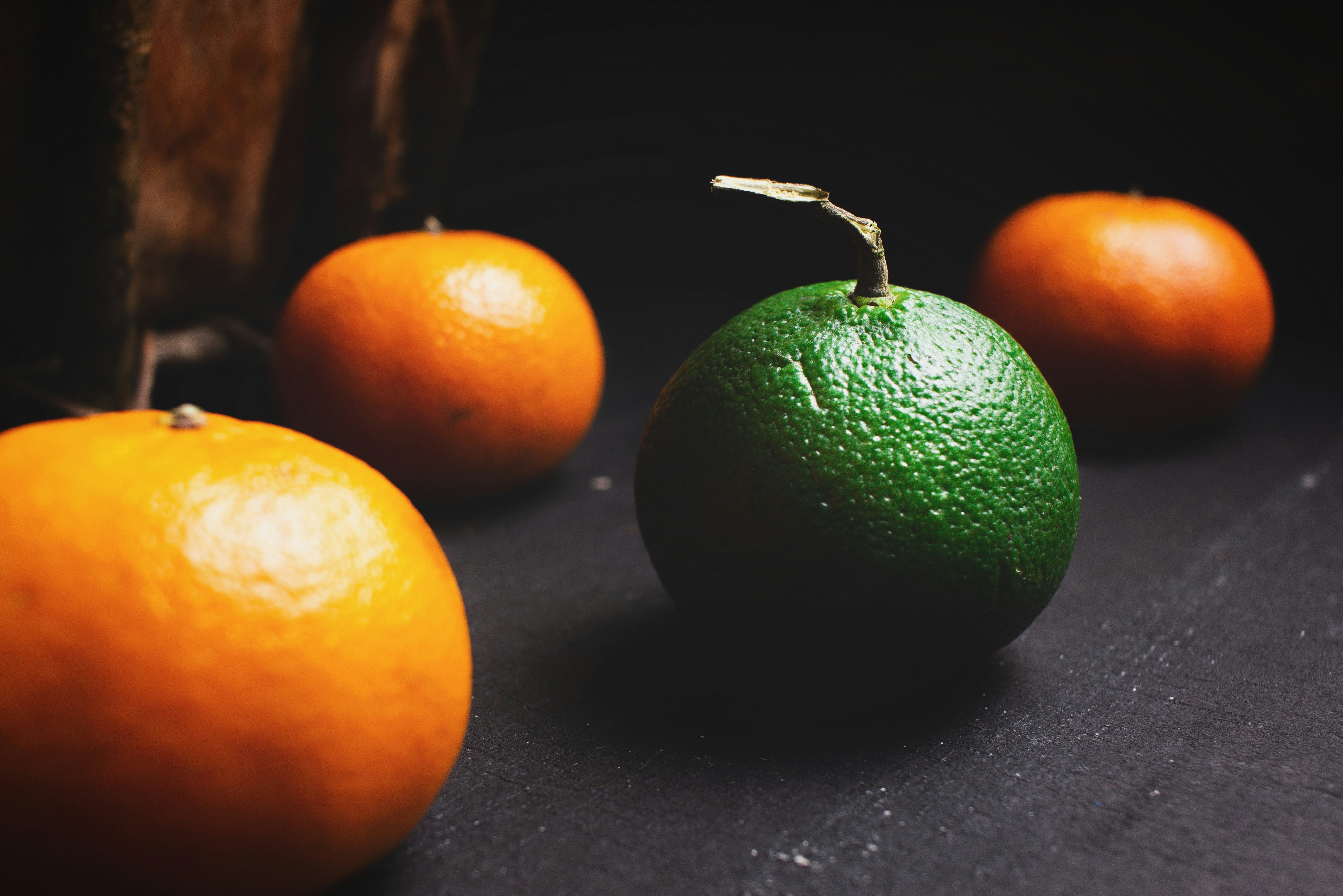 A vibrant green lime stands out among several bright oranges on a dark surface, highlighting the contrast in color and texture.