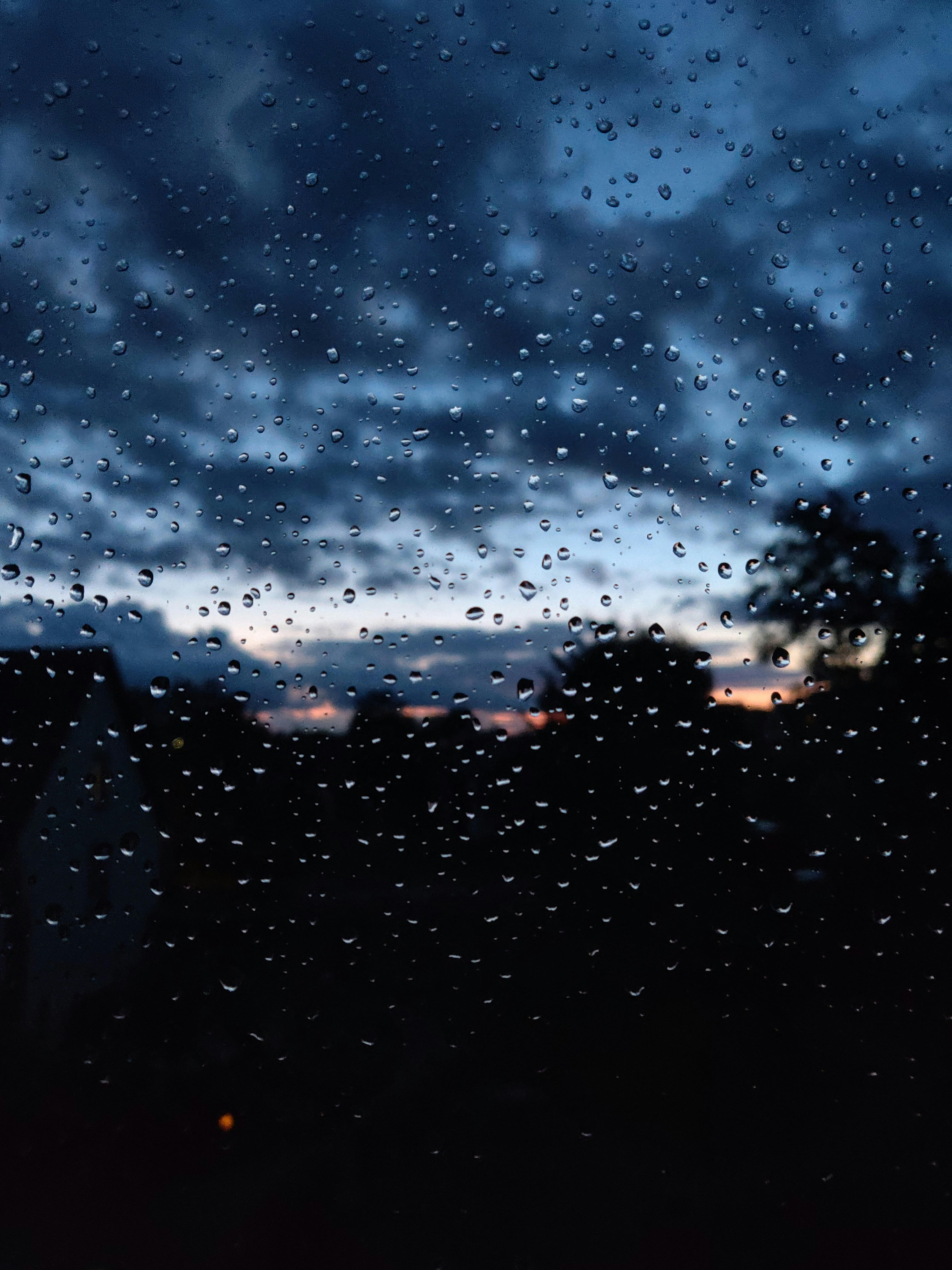 Close-up of raindrops on a windowpane, capturing the twilight sky with dark clouds and hints of color at dusk.