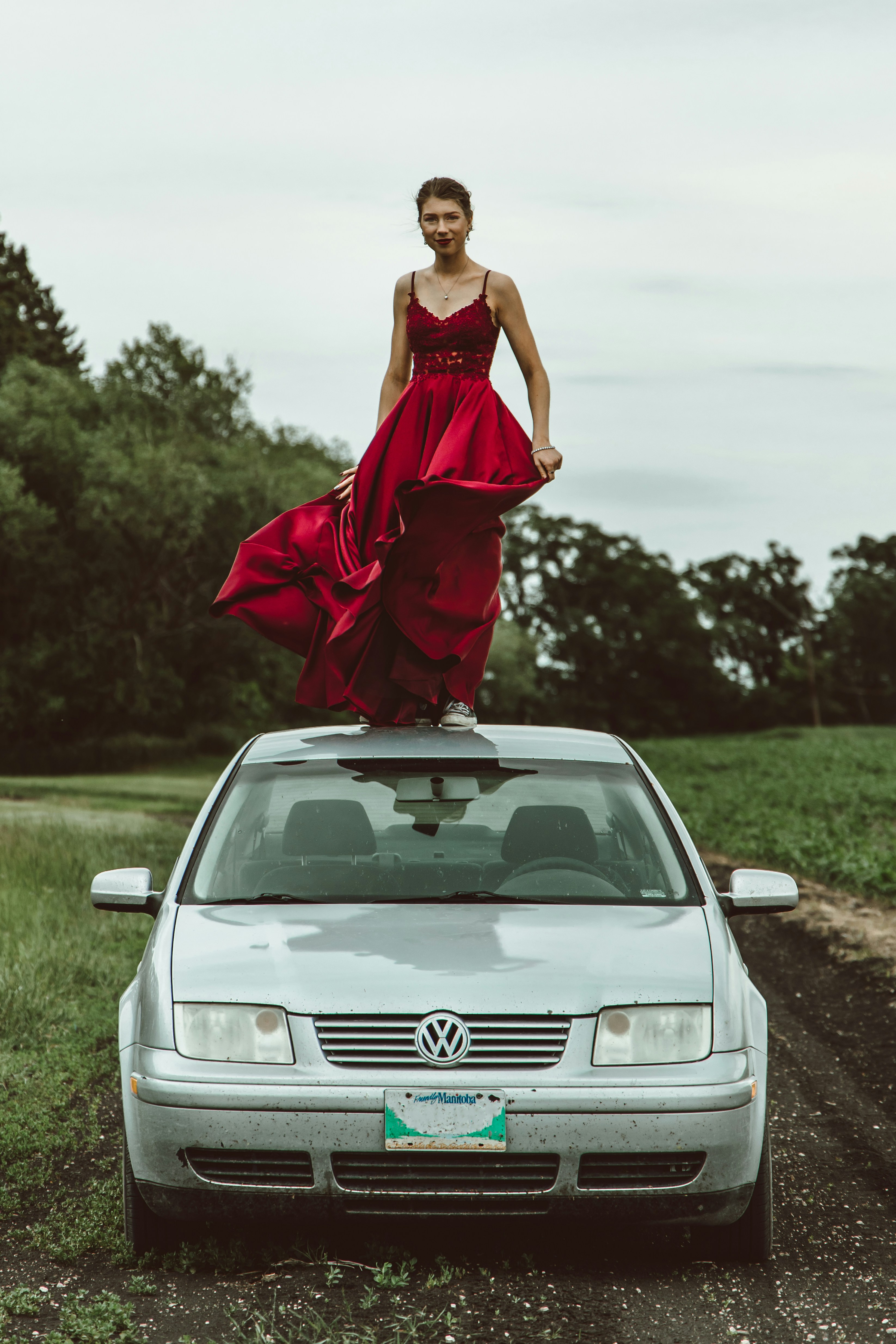 Model in a flowing red gown stands atop a silver Volkswagen, showcasing a blend of elegance and rebellion. The backdrop features lush greenery and an overcast sky.