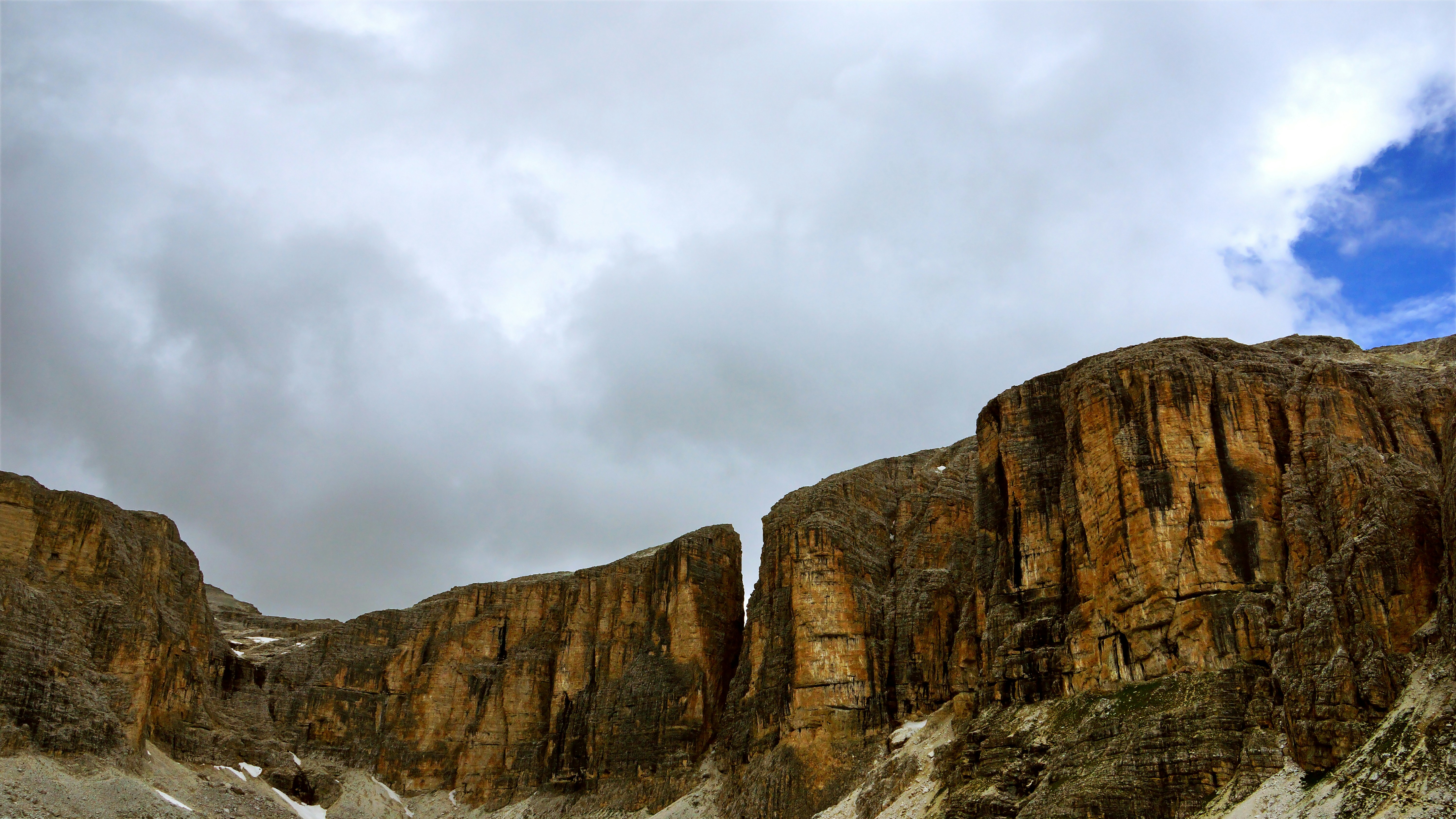 Towering cliffs rise dramatically against a backdrop of swirling clouds, showcasing the rugged beauty of the landscape. Snow patches linger on the ground, hinting at the harsh elements of the environment.