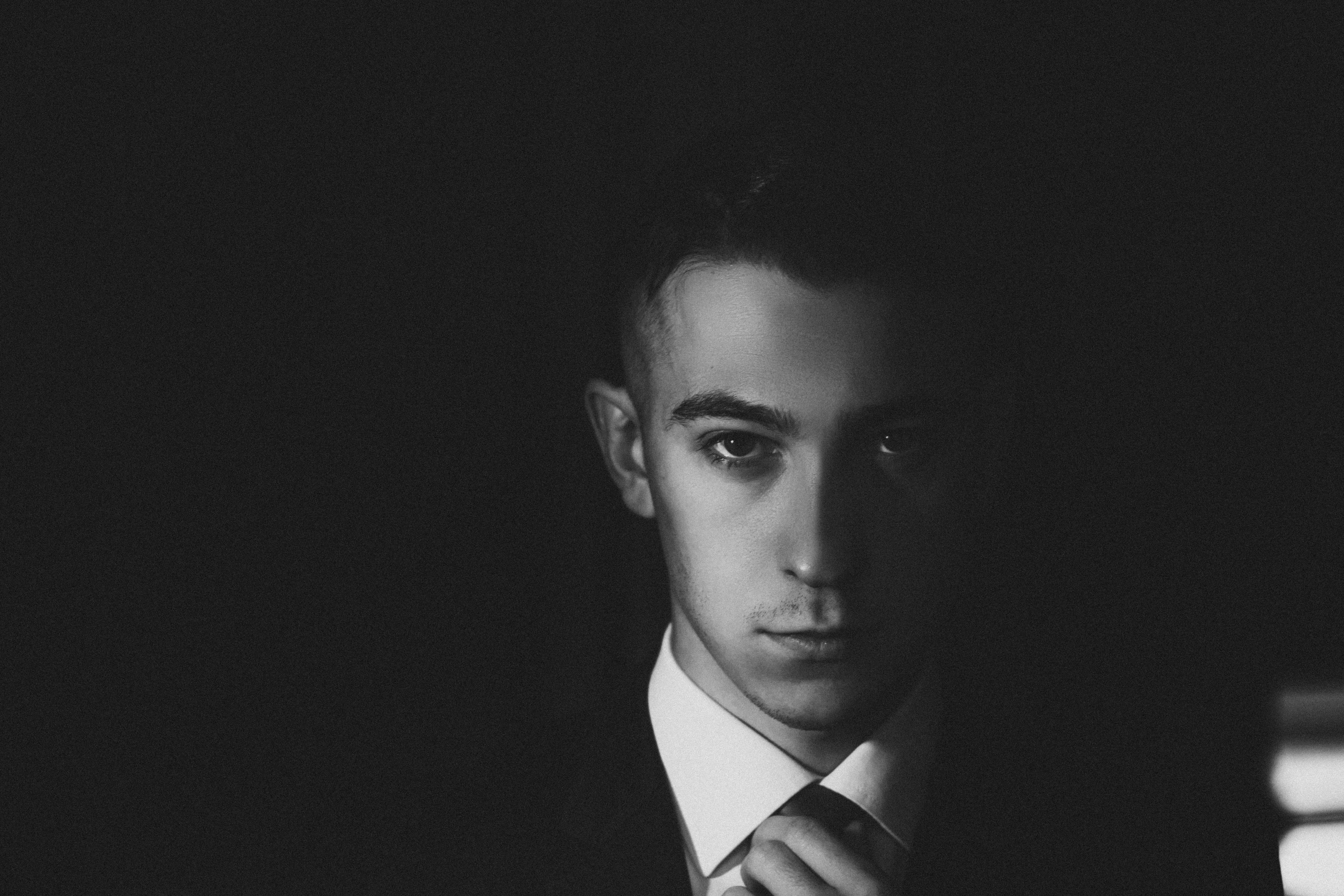 A young man in a suit gazes thoughtfully, partially illuminated against a dark backdrop, conveying a sense of depth and contemplation.