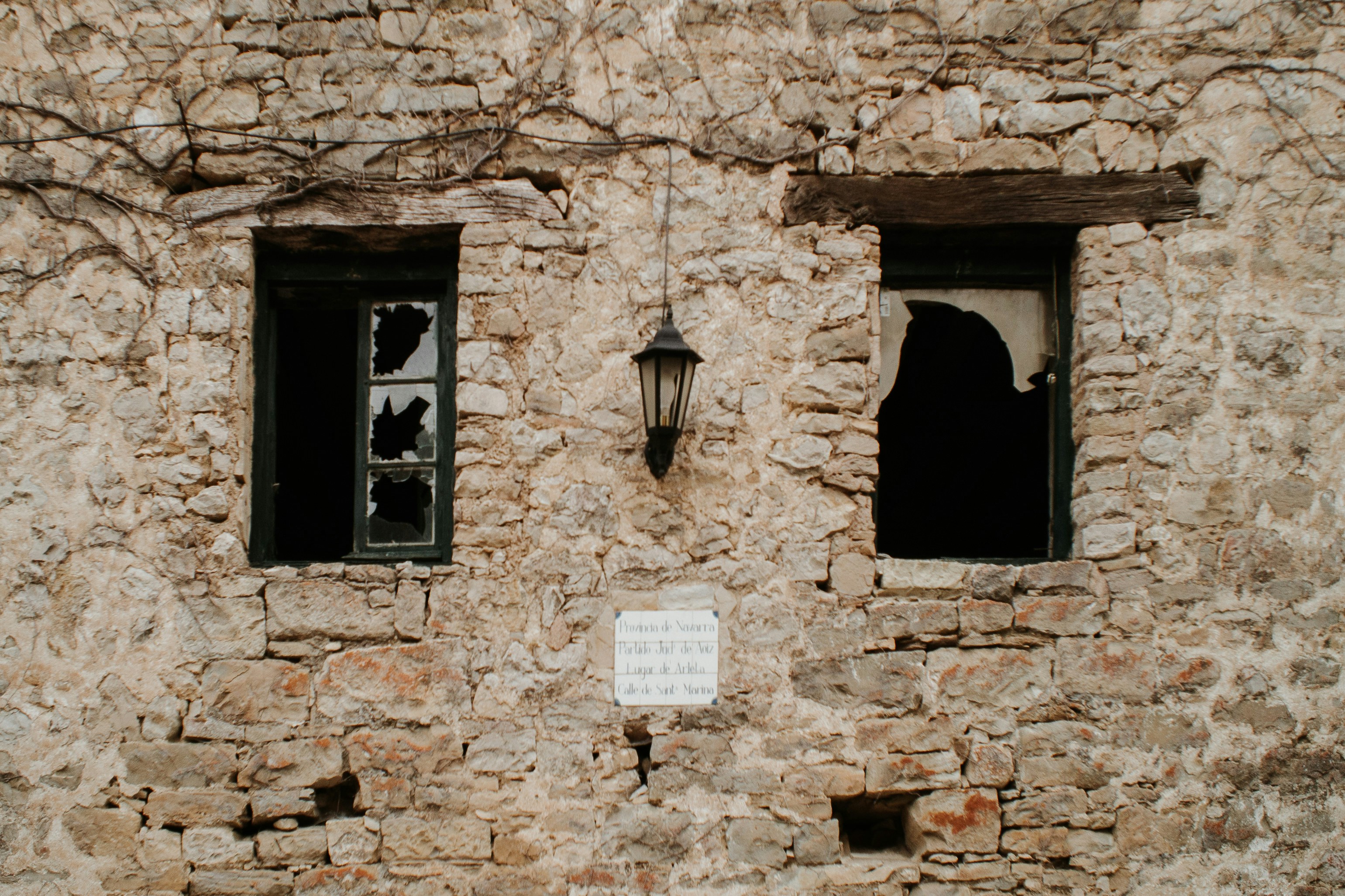 Weathered stone wall featuring two broken windows and a vintage lantern, accompanied by a faded sign. A glimpse into a bygone era.