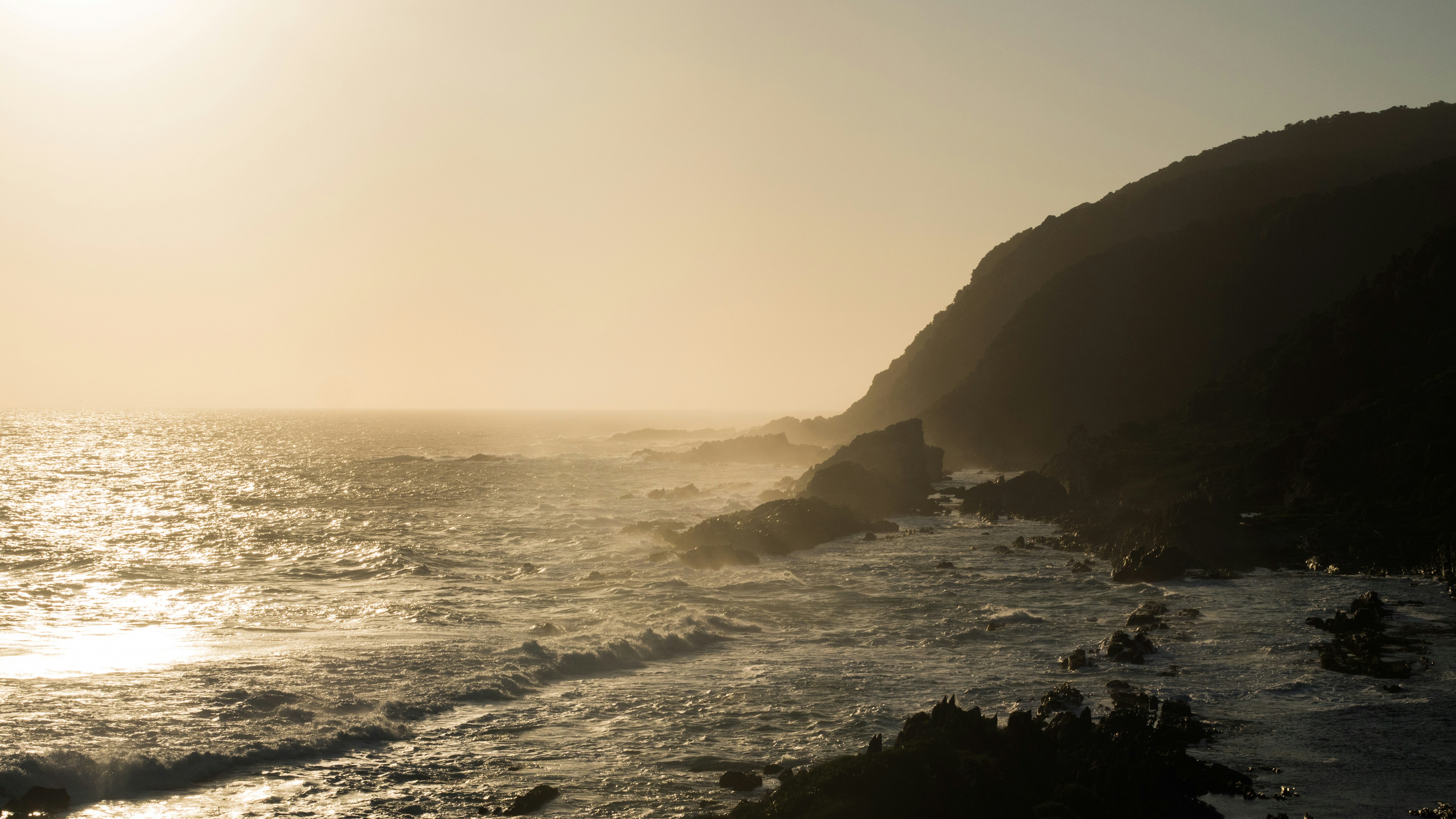 Sunlit ocean waves crash against a rocky cliff under a hazy sky.