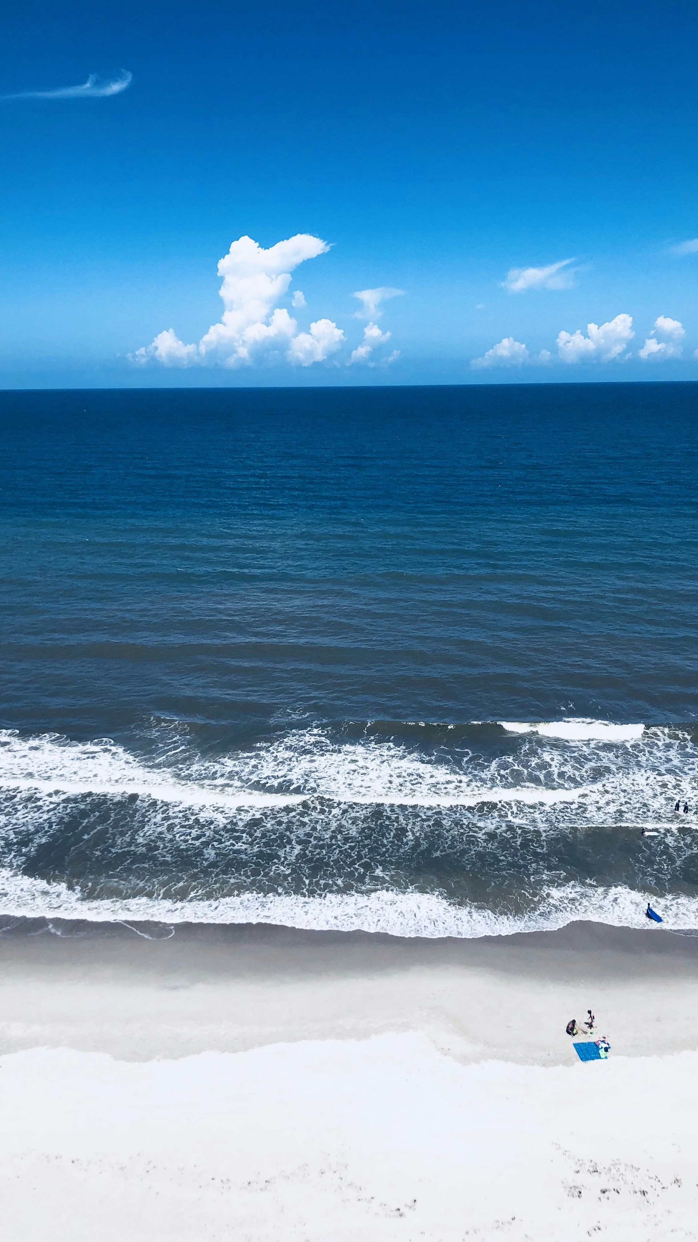 Gentle waves lapping against a sandy beach under a vibrant blue sky with scattered clouds. Two figures relax on the shore.