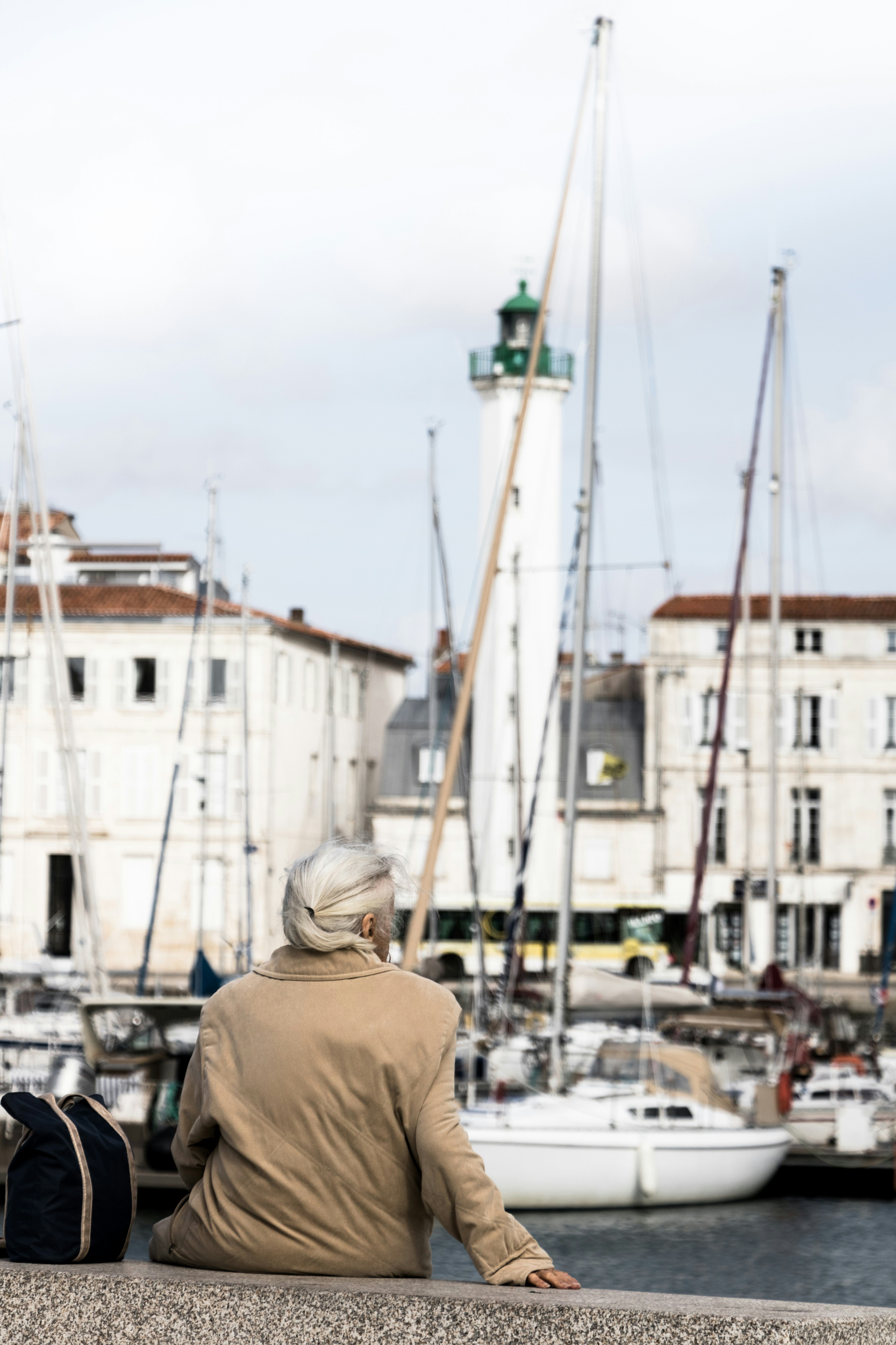 Elderly figure seated by a harbor, gazing at boats and a lighthouse in the background, conveying a sense of tranquility and nostalgia.