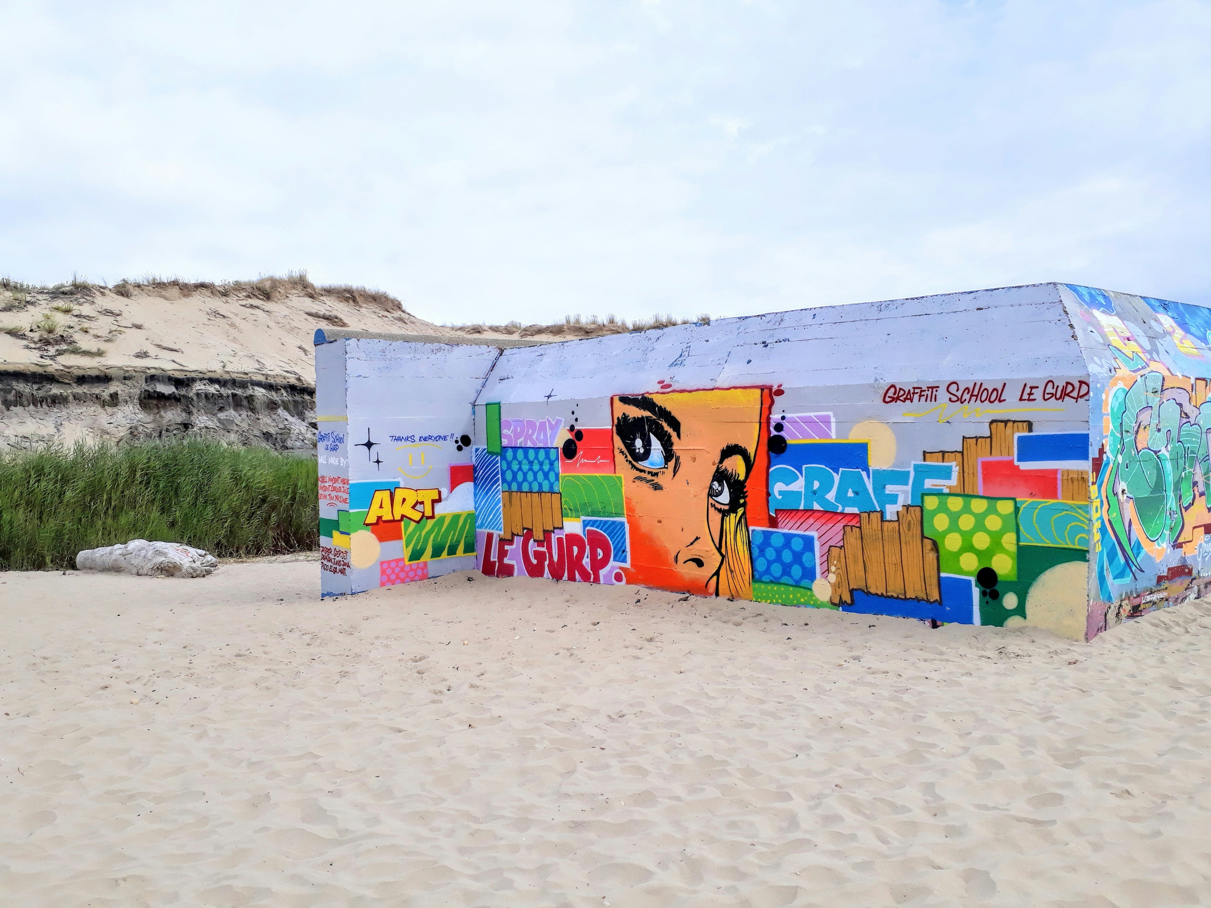 Colorful graffiti mural on a beachside wall, featuring artistic designs and a striking face illustration. The surrounding sandy landscape enhances the vibrant colors.