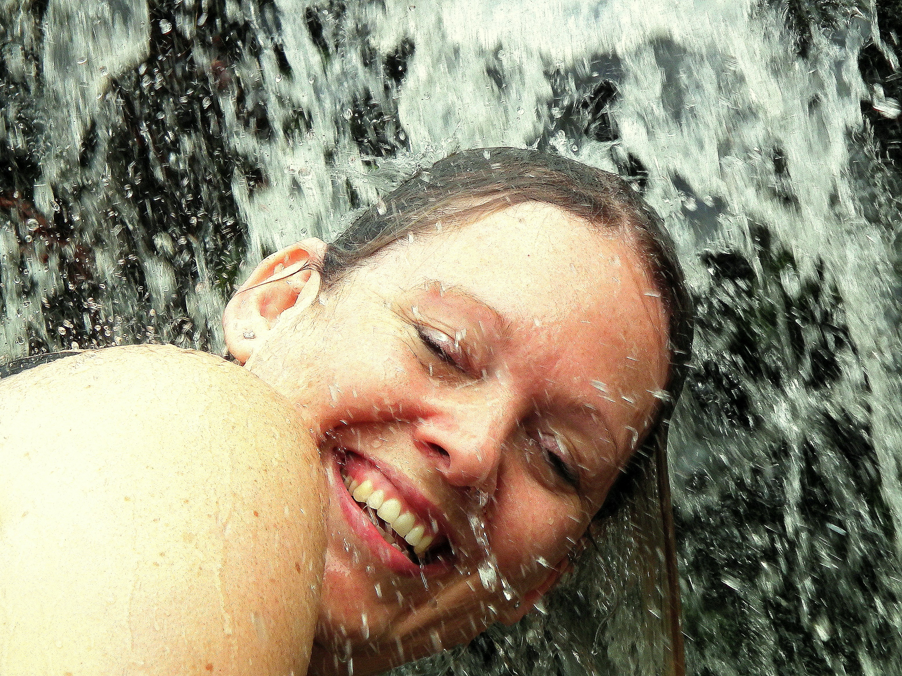 Close-up photograph of a smiling woman beneath a curtain of water, capturing droplets in motion.