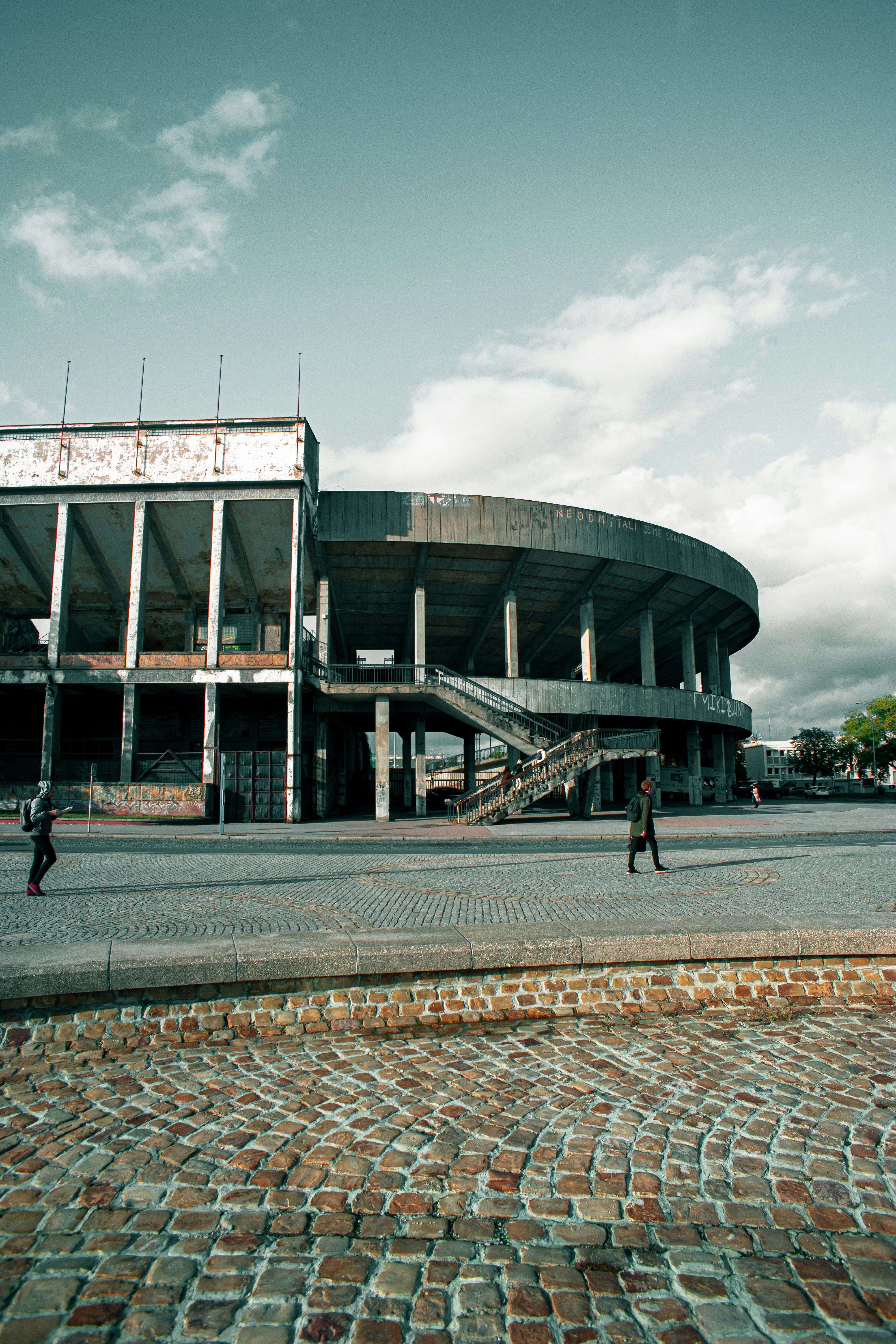 Abandoned concrete stadium with a spiral staircase, surrounded by cobblestone pavement and a cloudy sky. The scene captures urban decay and the passage of time.