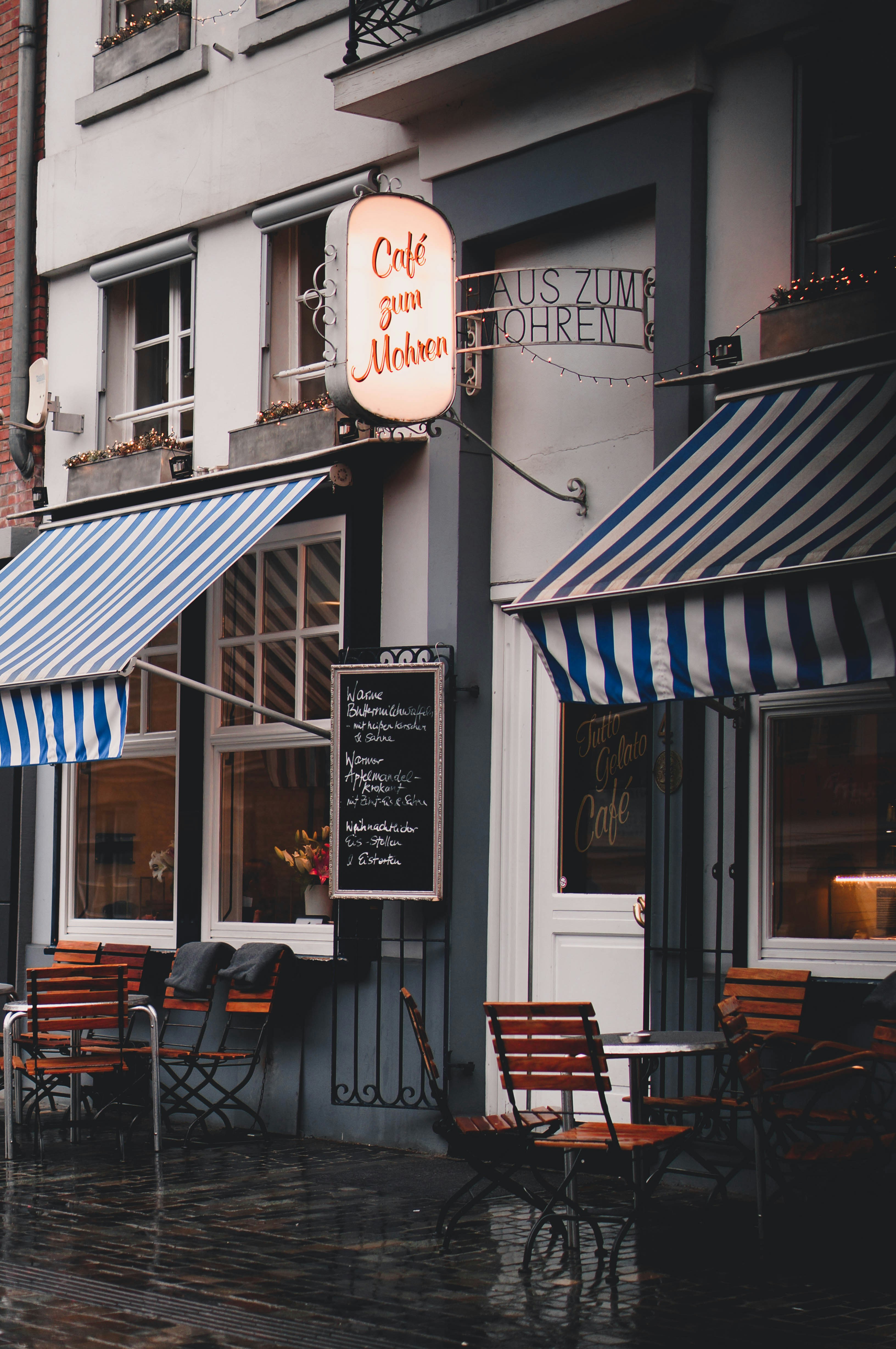 Charming café facade adorned with striped awnings and a glowing sign, inviting patrons to enjoy a warm drink. A menu board adds a touch of local flavor.