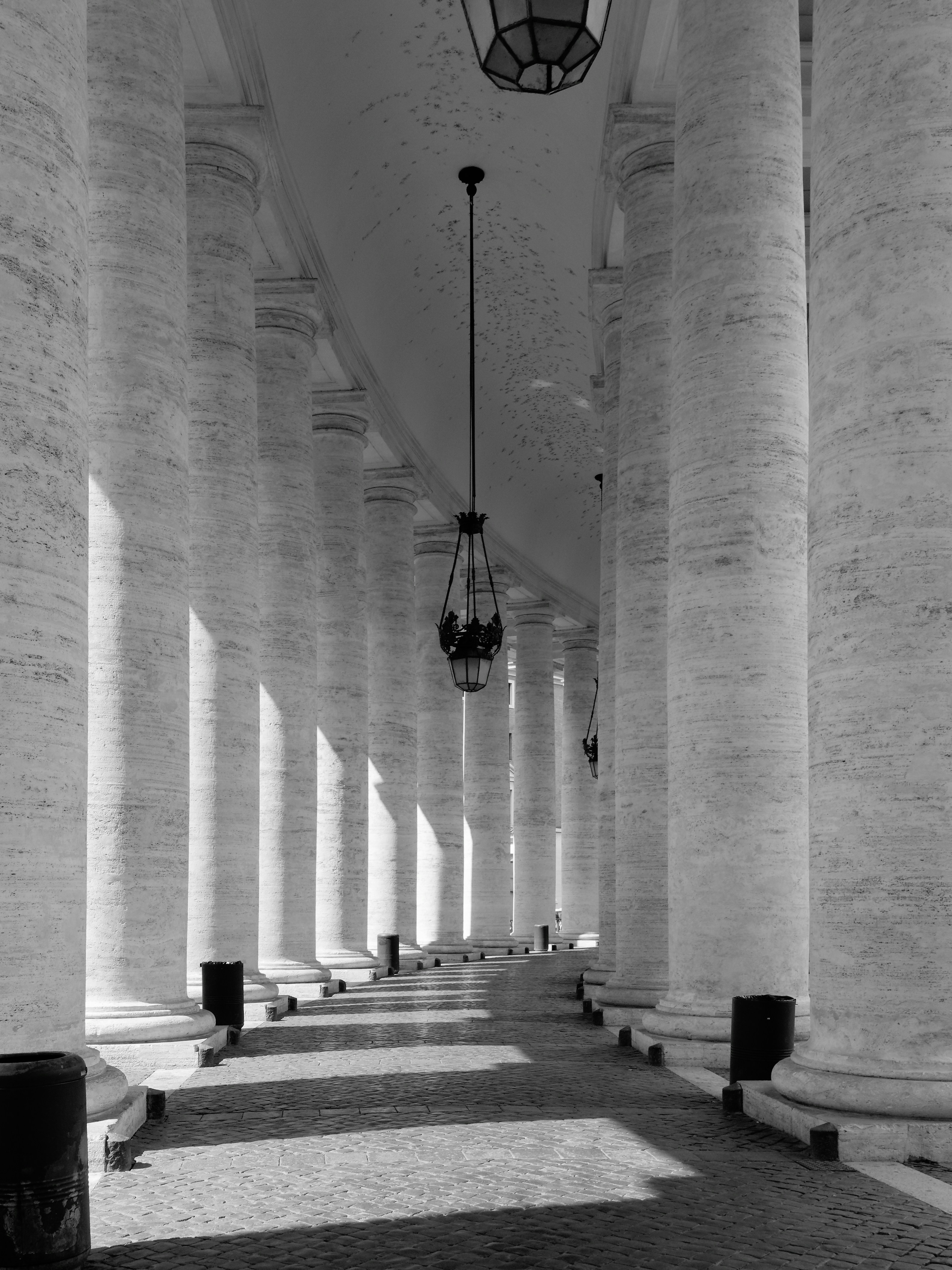 Black-and-white architectural photograph of a long colonnade lined with tall pillars and hanging lanterns, leading the eye toward a distant vanishing point.