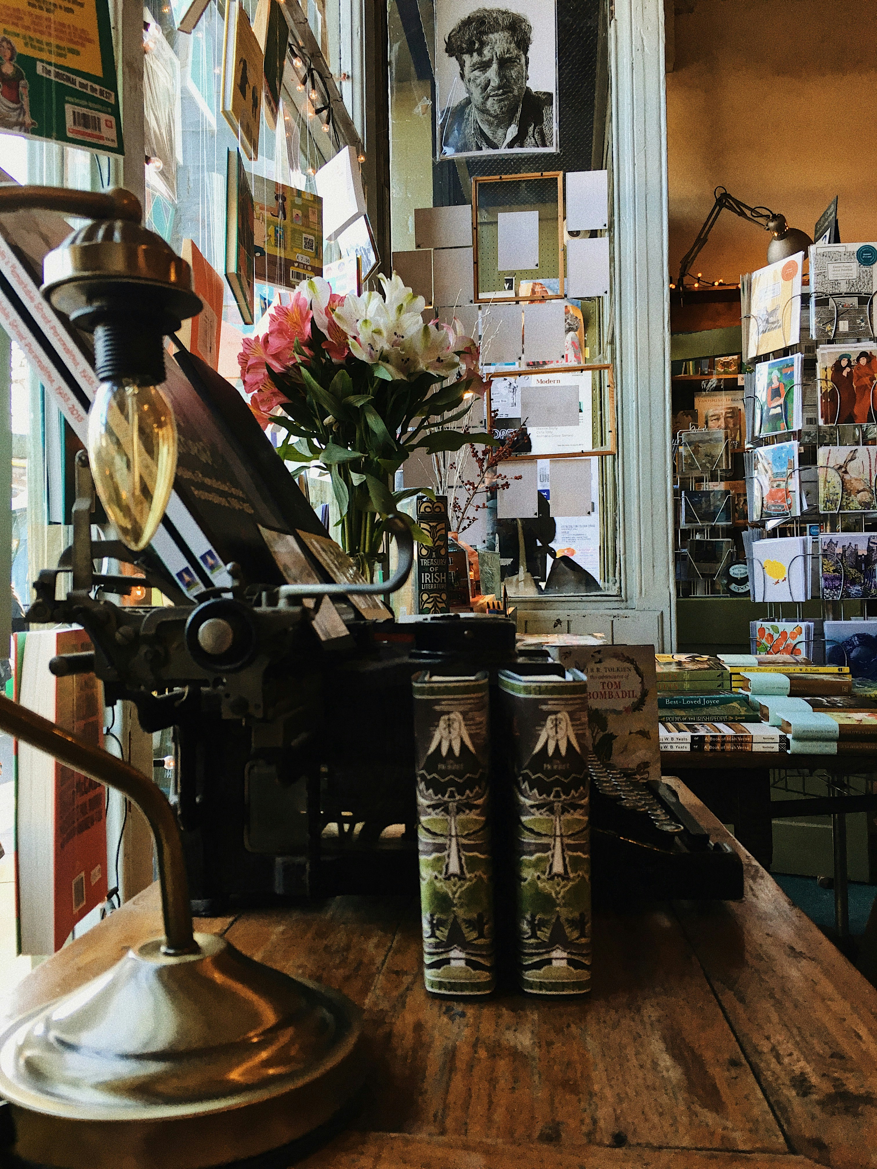 A vintage printing press sits beside a vase of flowers, surrounded by colorful cards and books in a quaint shop.