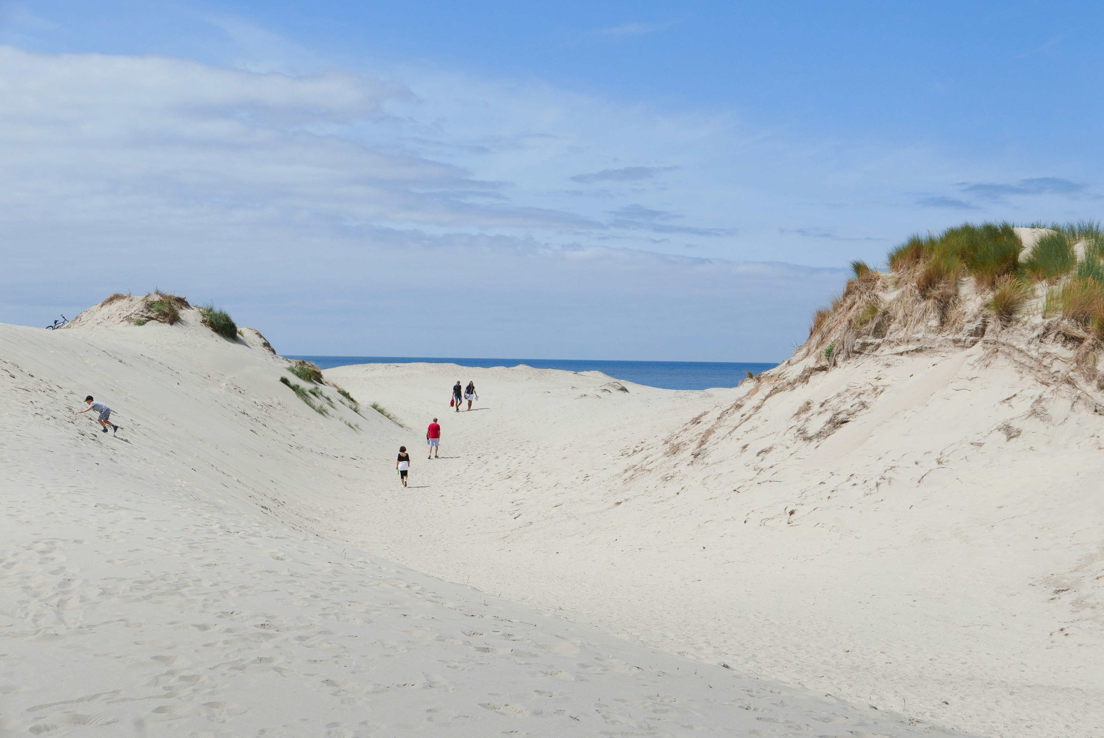 A scenic view of sandy dunes leading to the ocean, with people strolling along the paths under a partly cloudy sky.